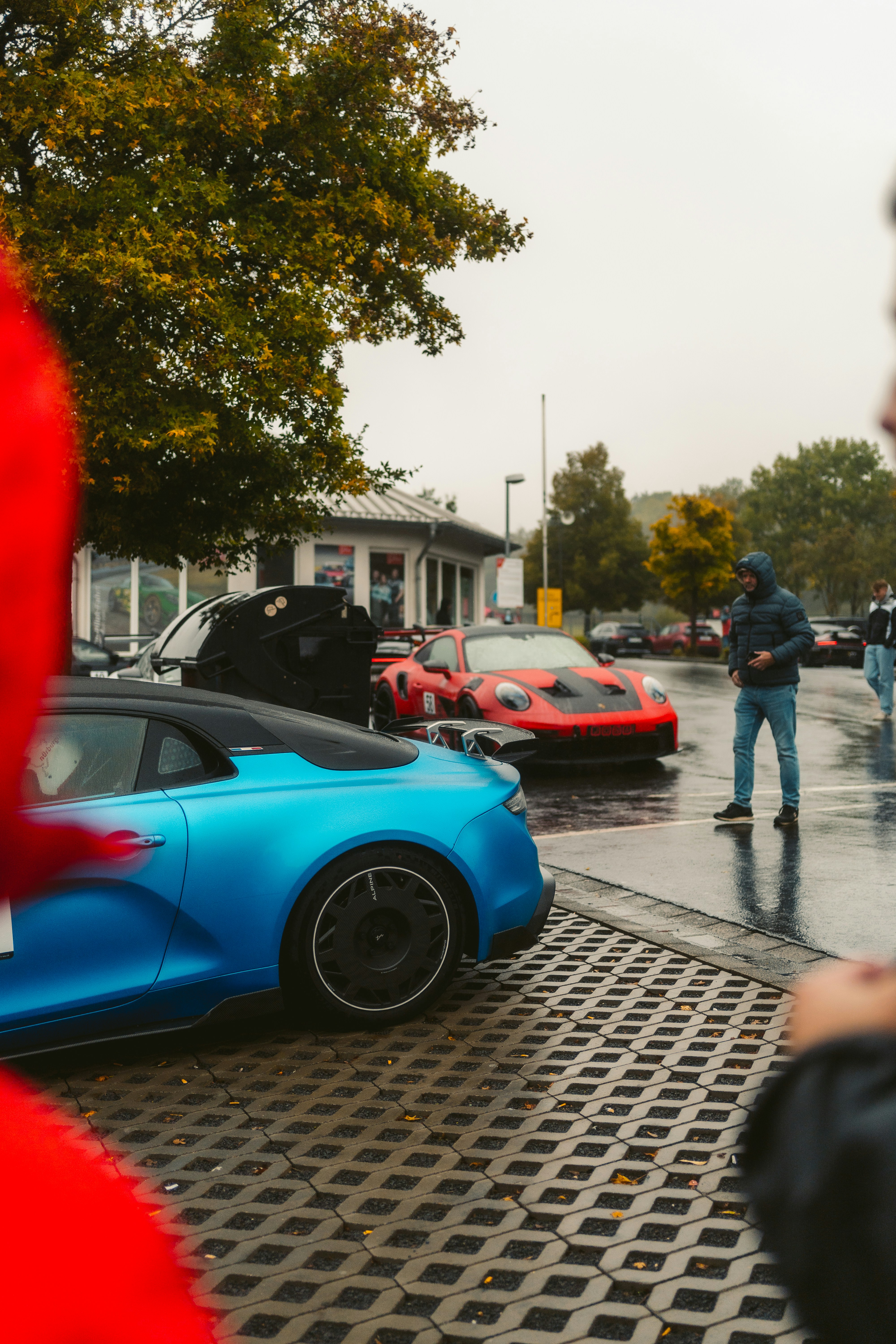 A vibrant blue sports car stands prominently in a rainy parking lot, with a red sports car and spectators in the background. The scene captures the essence of automotive passion amidst drizzly weather.