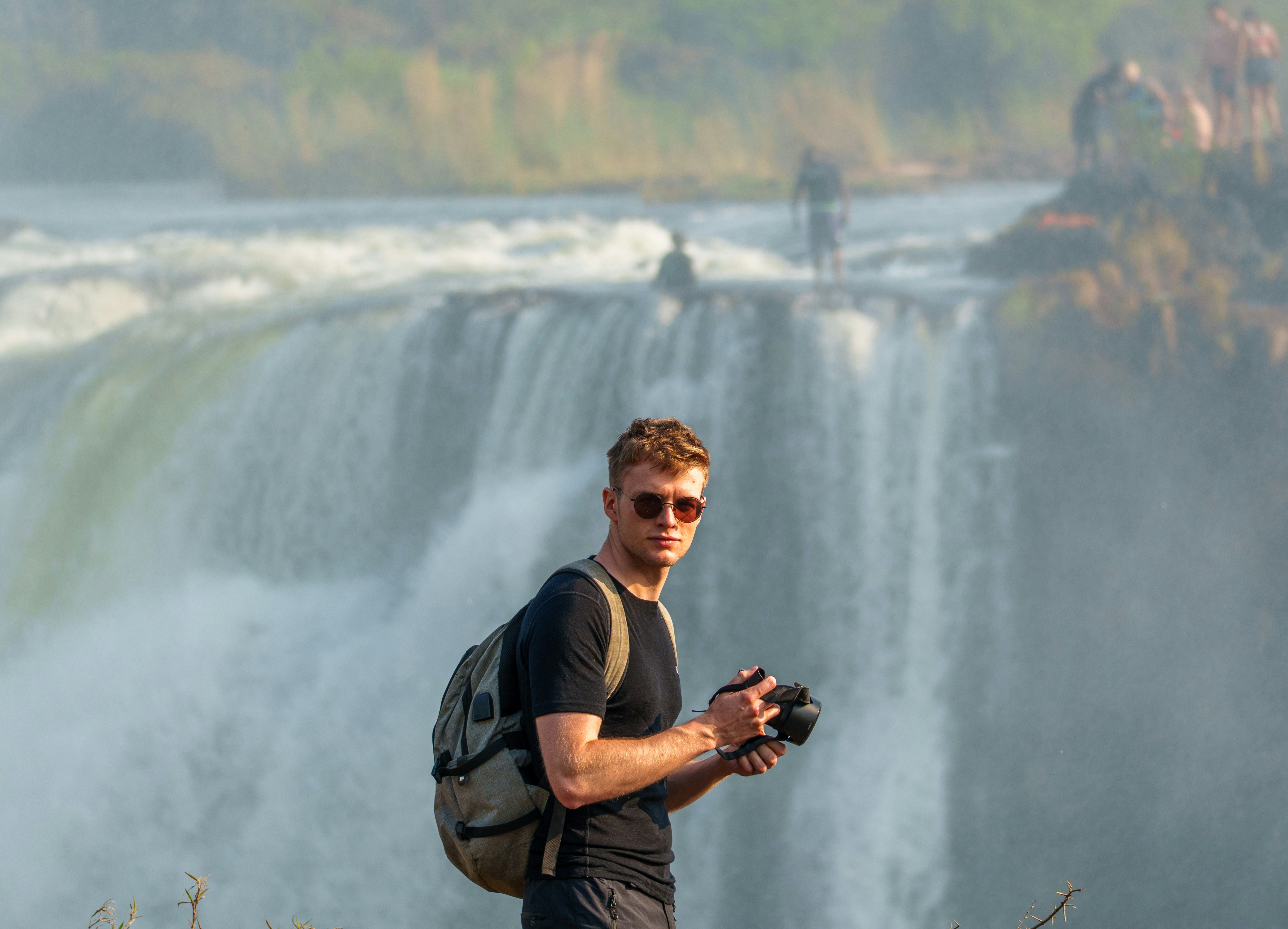 A man stands in front of victoria falls with a camera. | Man with camera and backpack near waterfall