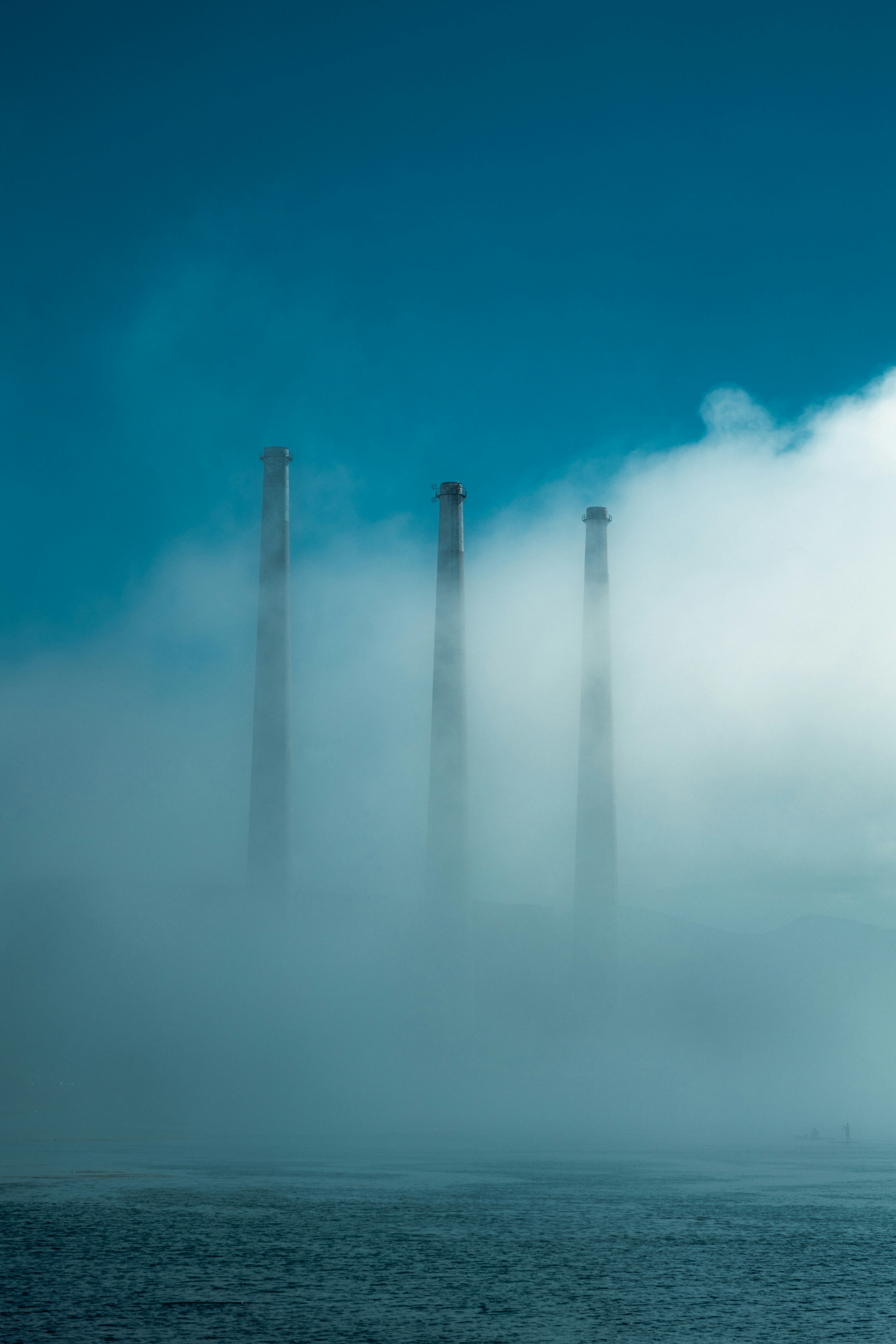 Three smokestacks emerge from thick fog over water.