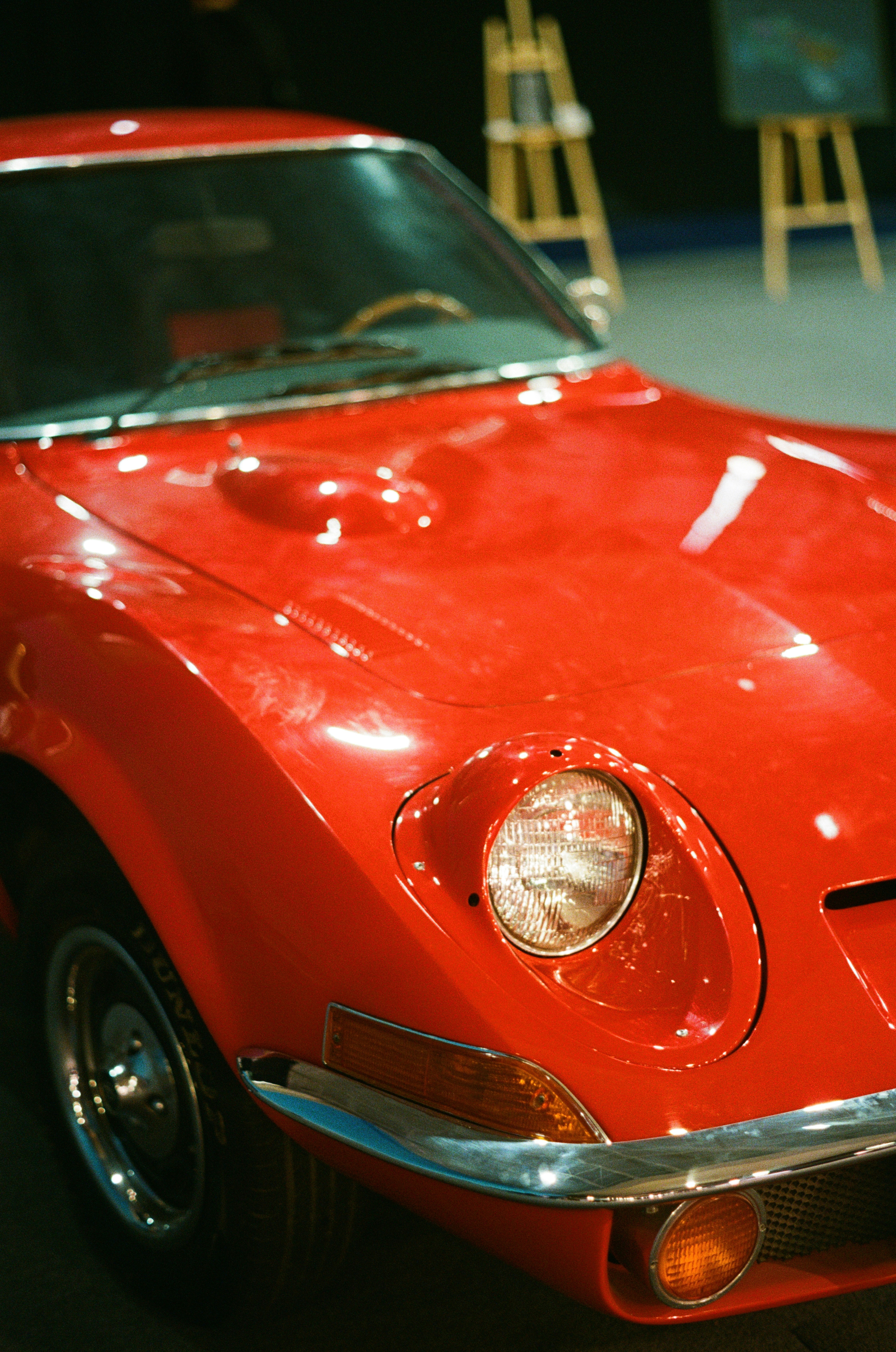 Close-up of a vintage red sports car showcasing its sleek curves and detailed headlights. The setting hints at an exhibition atmosphere.