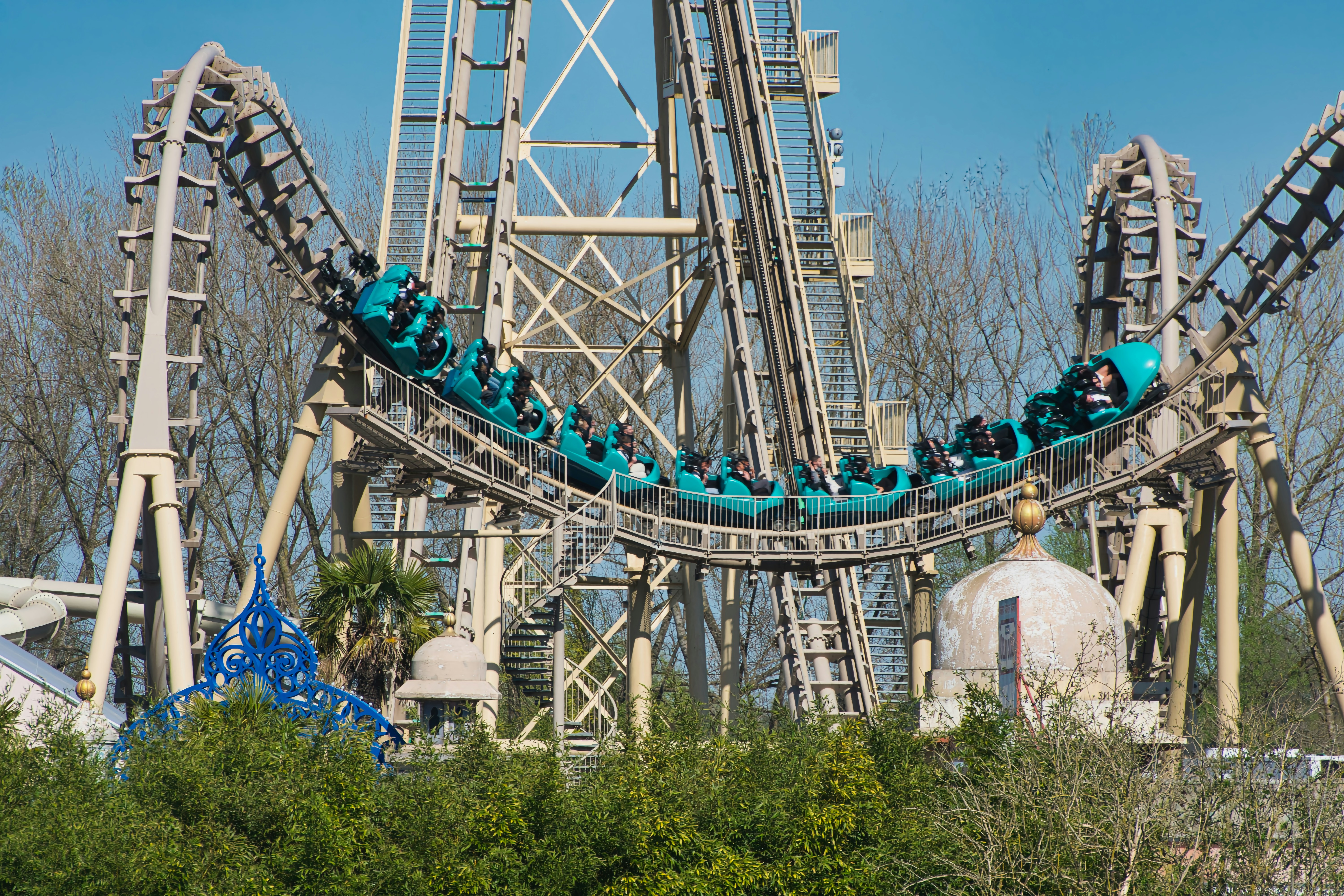 Steel Serpent in Flight: Teal-Seat Roller Coaster Arcs Across a Sunlit Park