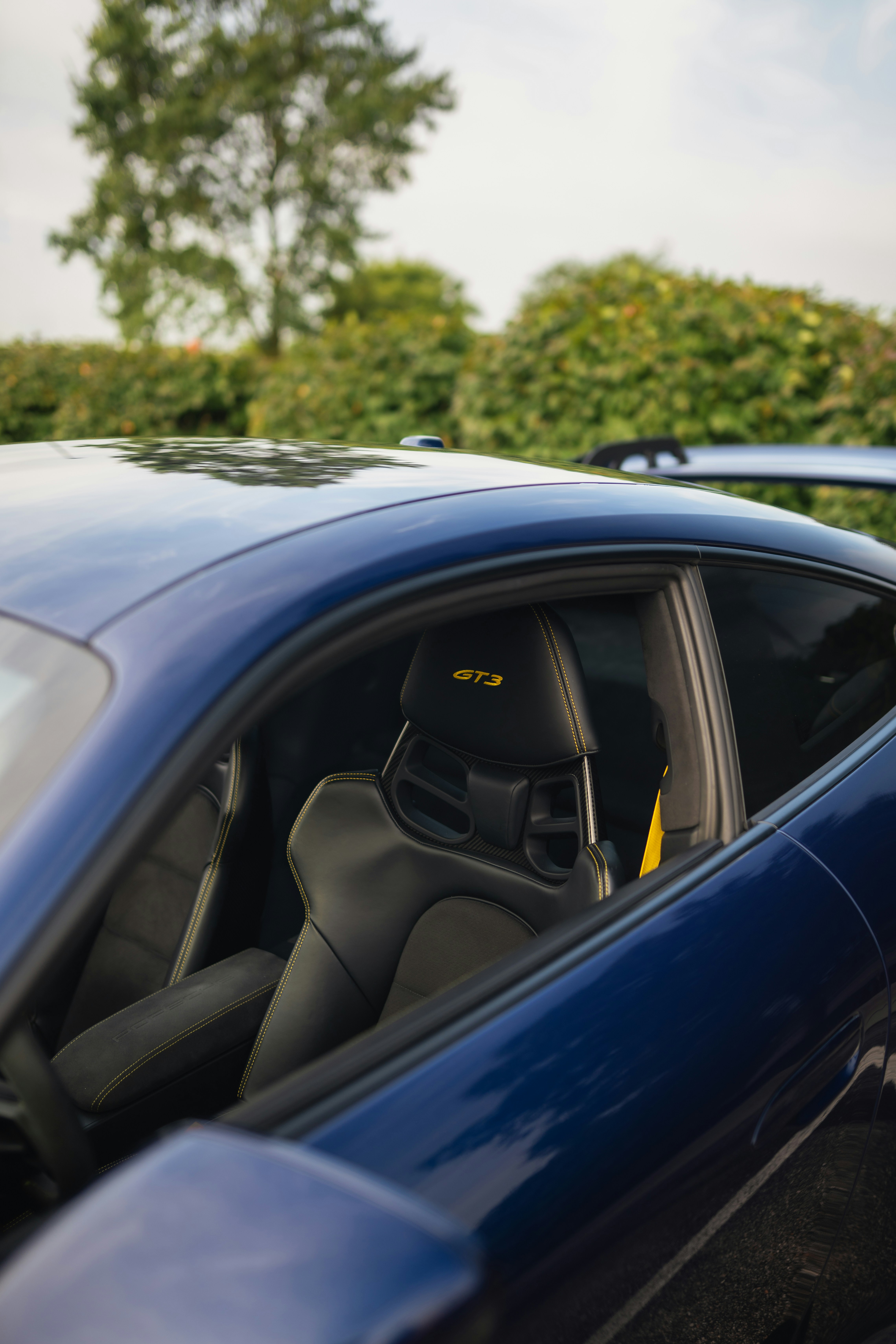 Interior view of a Porsche GT3 showcasing a sleek racing seat with yellow accents, framed by the car's blue exterior. The lush greenery in the background adds a vibrant contrast.