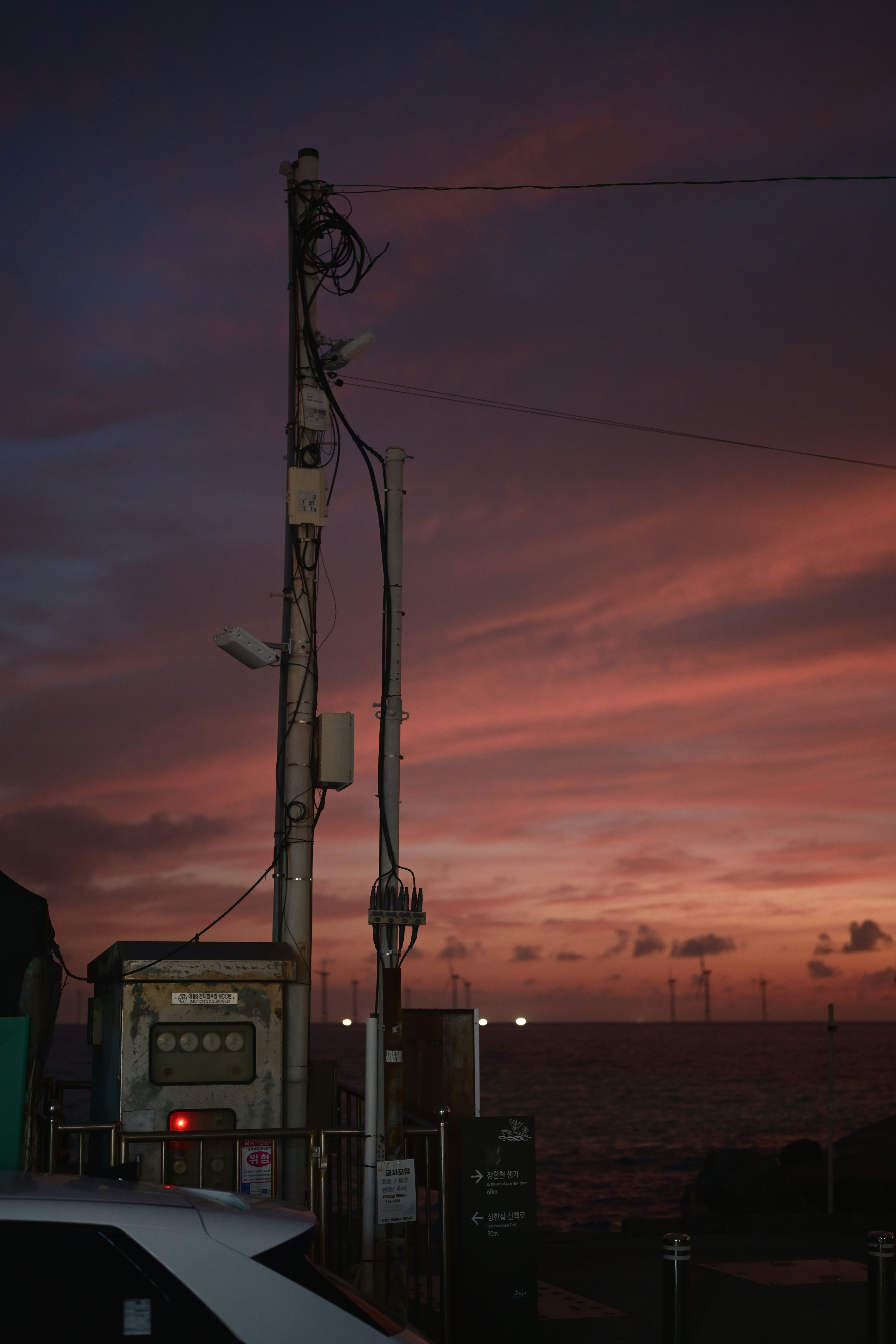 Silhouetted utility poles against a vibrant sunset, with wind turbines visible in the distance. The scene captures the intersection of technology and nature.