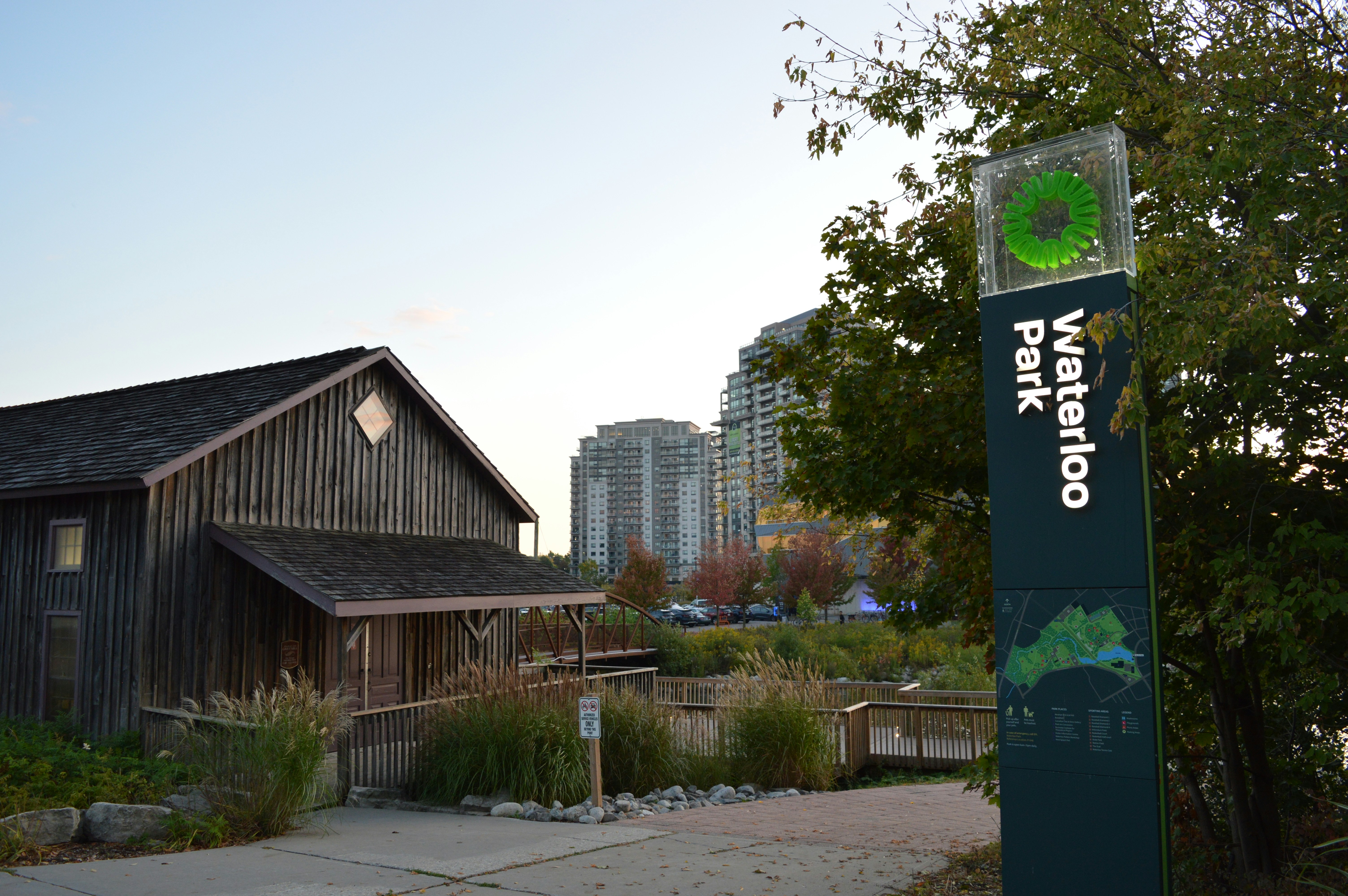 A rustic wooden building stands beside a modern park sign in Waterloo Park, surrounded by greenery and urban skyline.