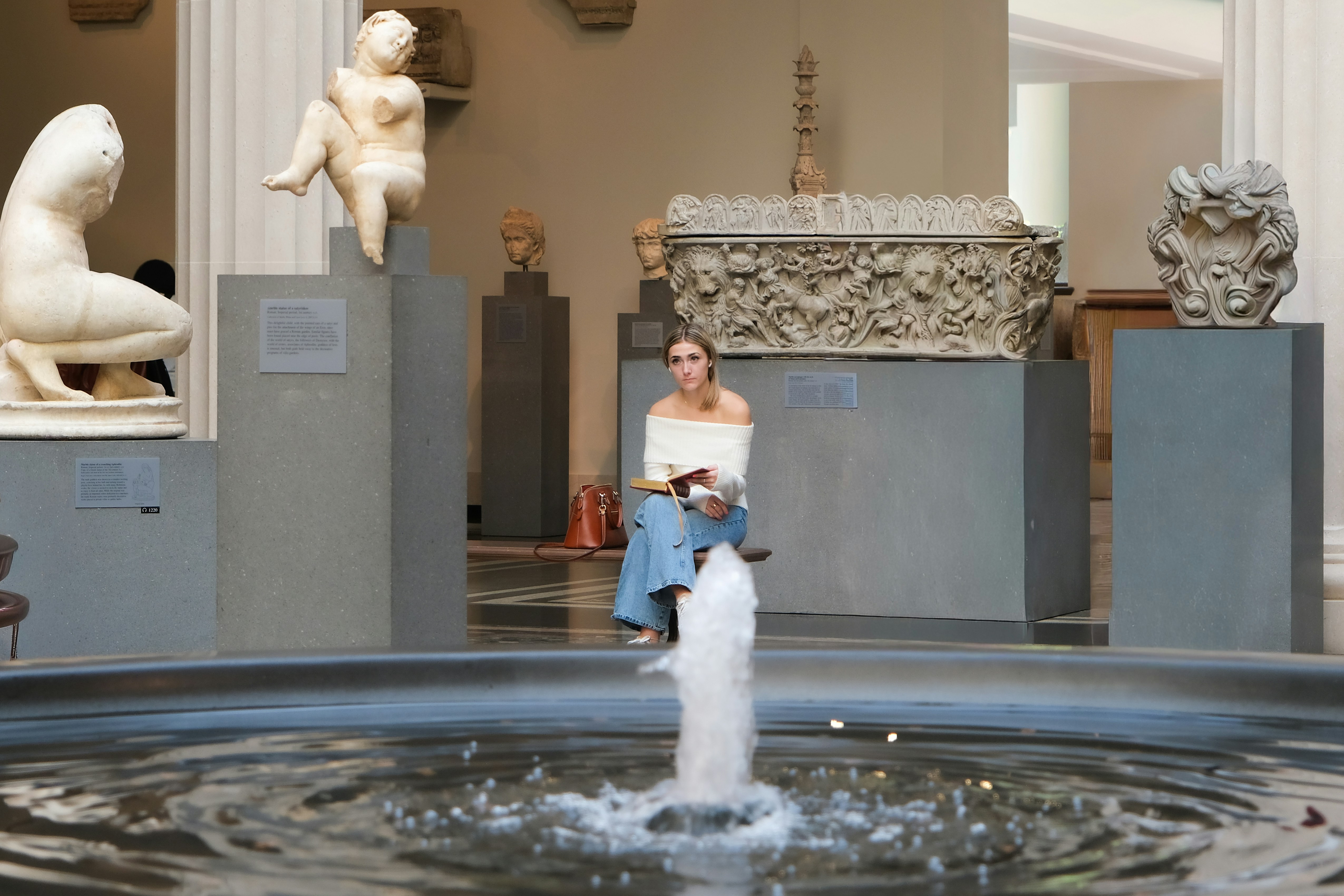 Woman sits by fountain in museum with sculptures.