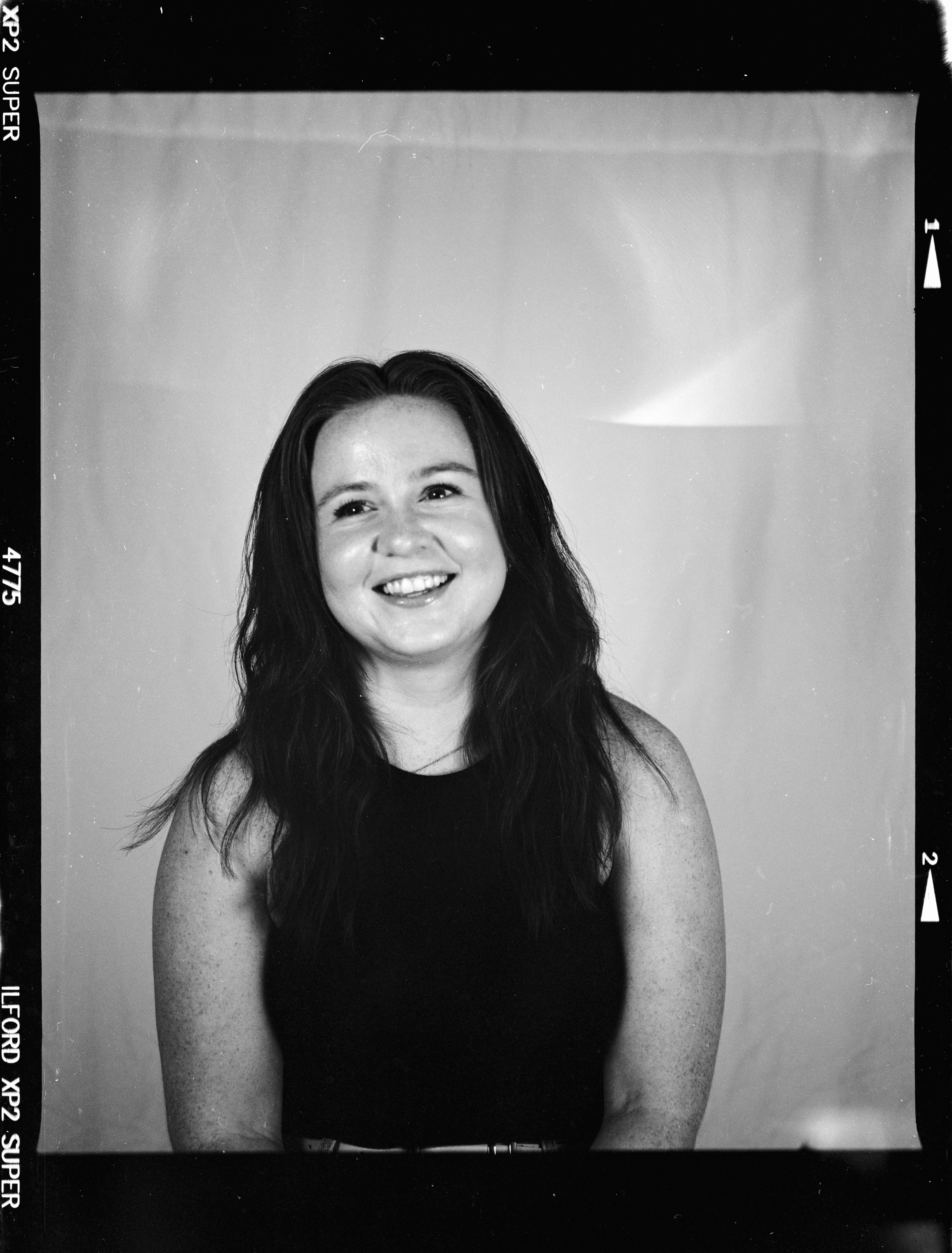 A woman with long hair smiles warmly against a softly lit backdrop, capturing a moment of genuine happiness. The photograph showcases a classic black and white aesthetic.