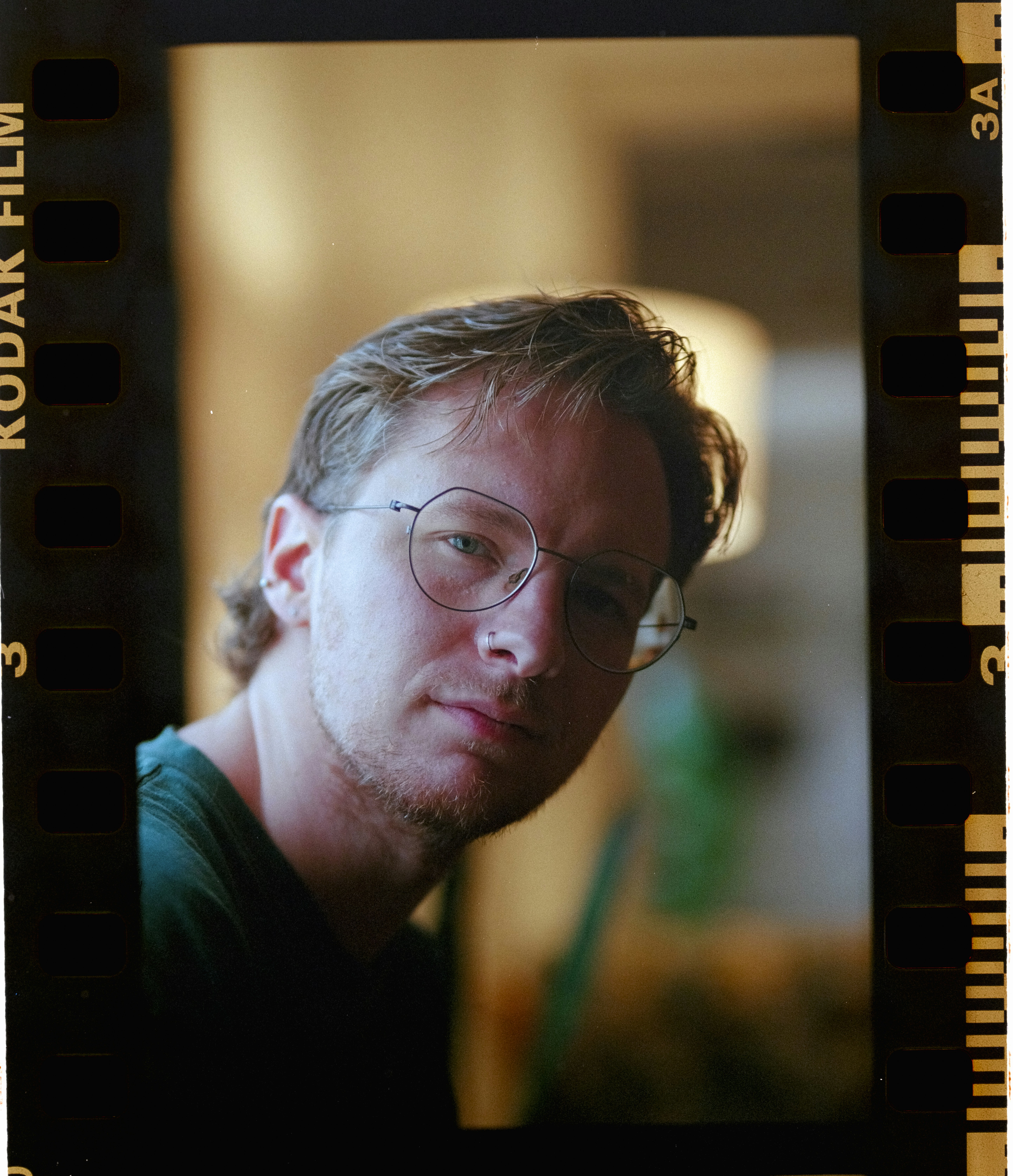 A young man with glasses gazes thoughtfully, framed by a film strip, with soft background bokeh suggesting a cozy indoor setting.