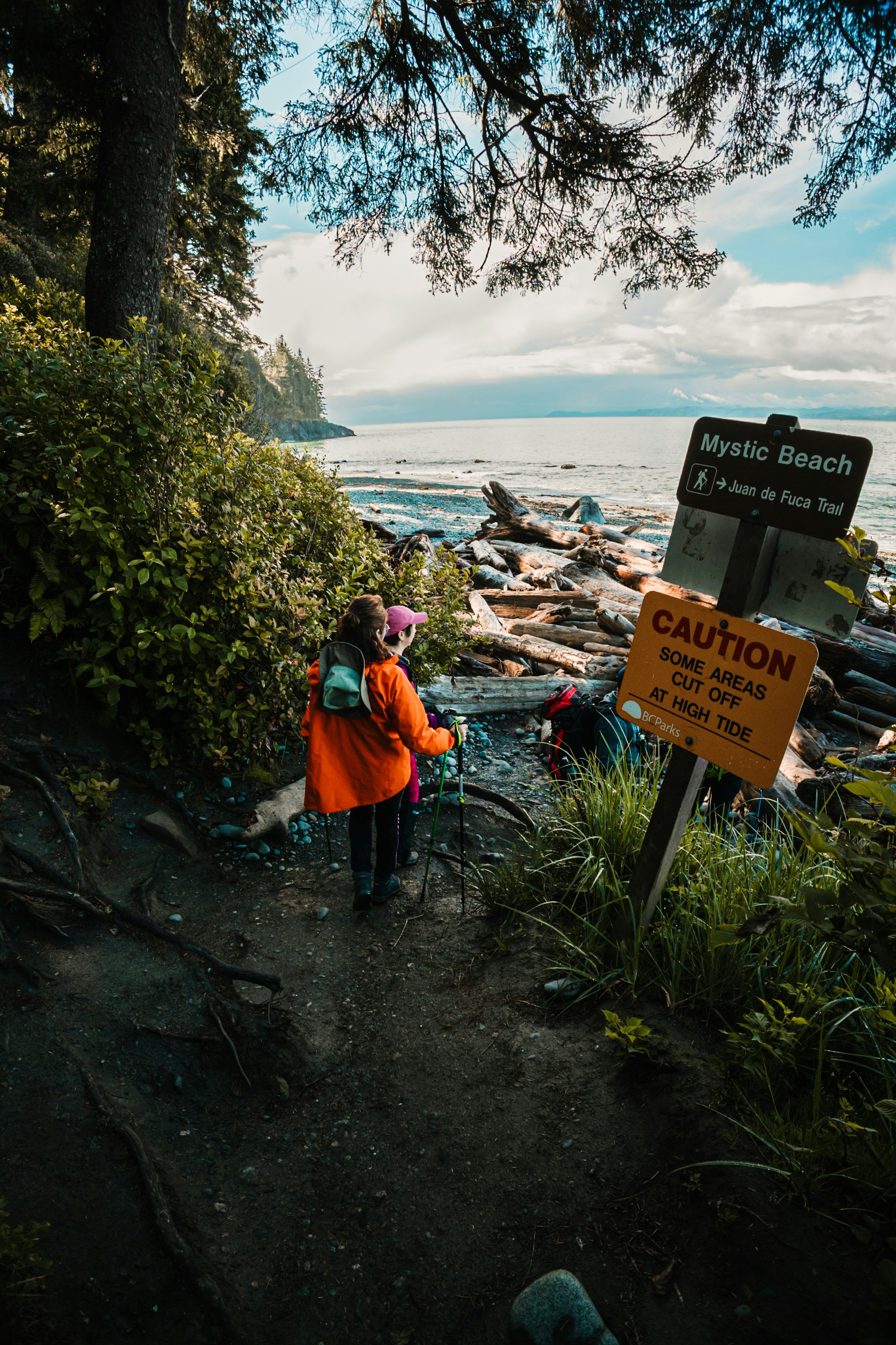 Hikers approach Mystic Beach, navigating through lush greenery and logs, with caution signs marking the trail. The serene ocean view beckons in the distance.