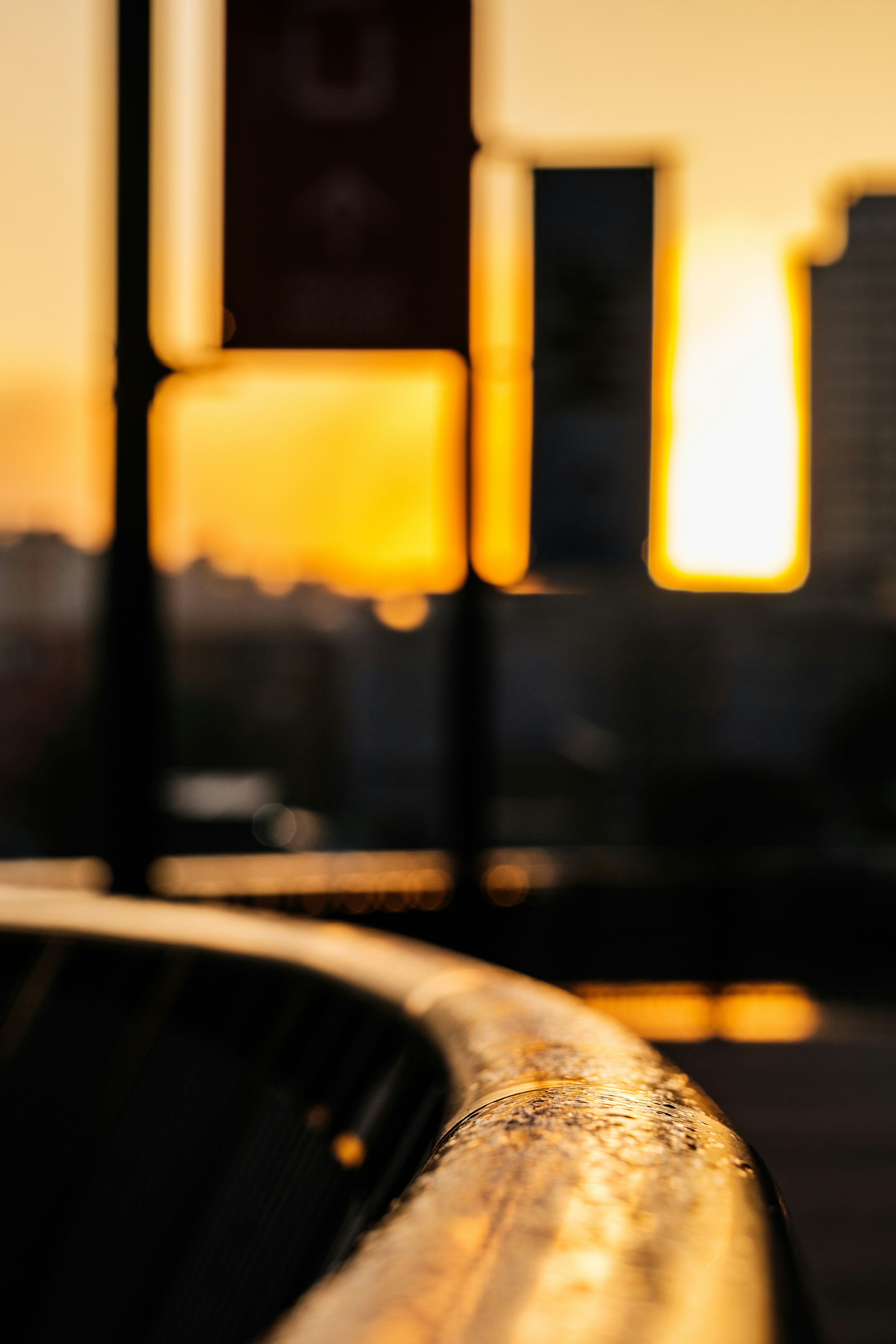Close-up of a wet railing reflecting the warm hues of a sunset, with blurred city banners in the background. The scene captures the tranquil beauty of urban twilight.