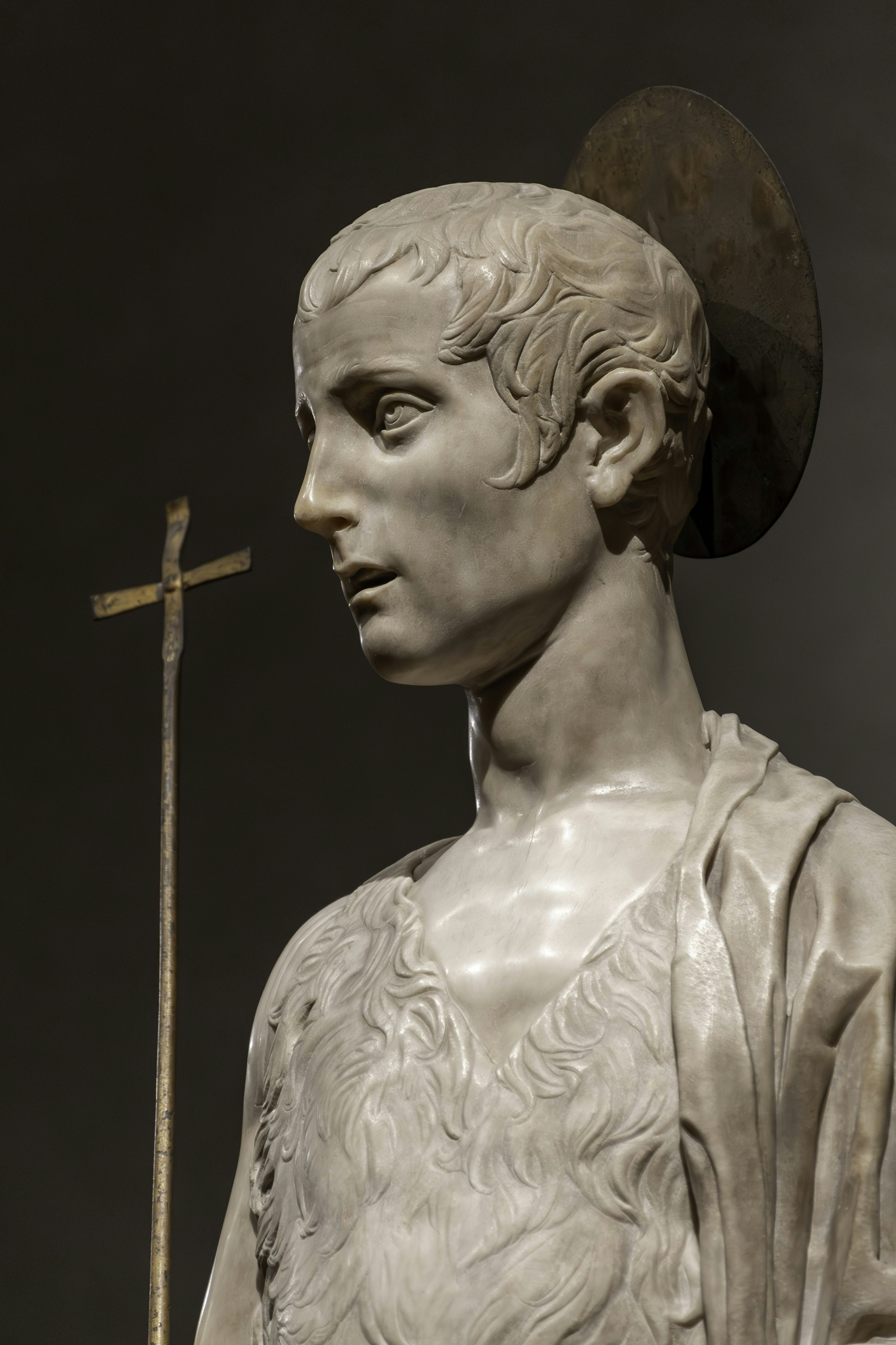 Detailed marble sculpture of a saint, featuring intricate textures and a serene expression, holding a cross. The soft lighting accentuates the craftsmanship.