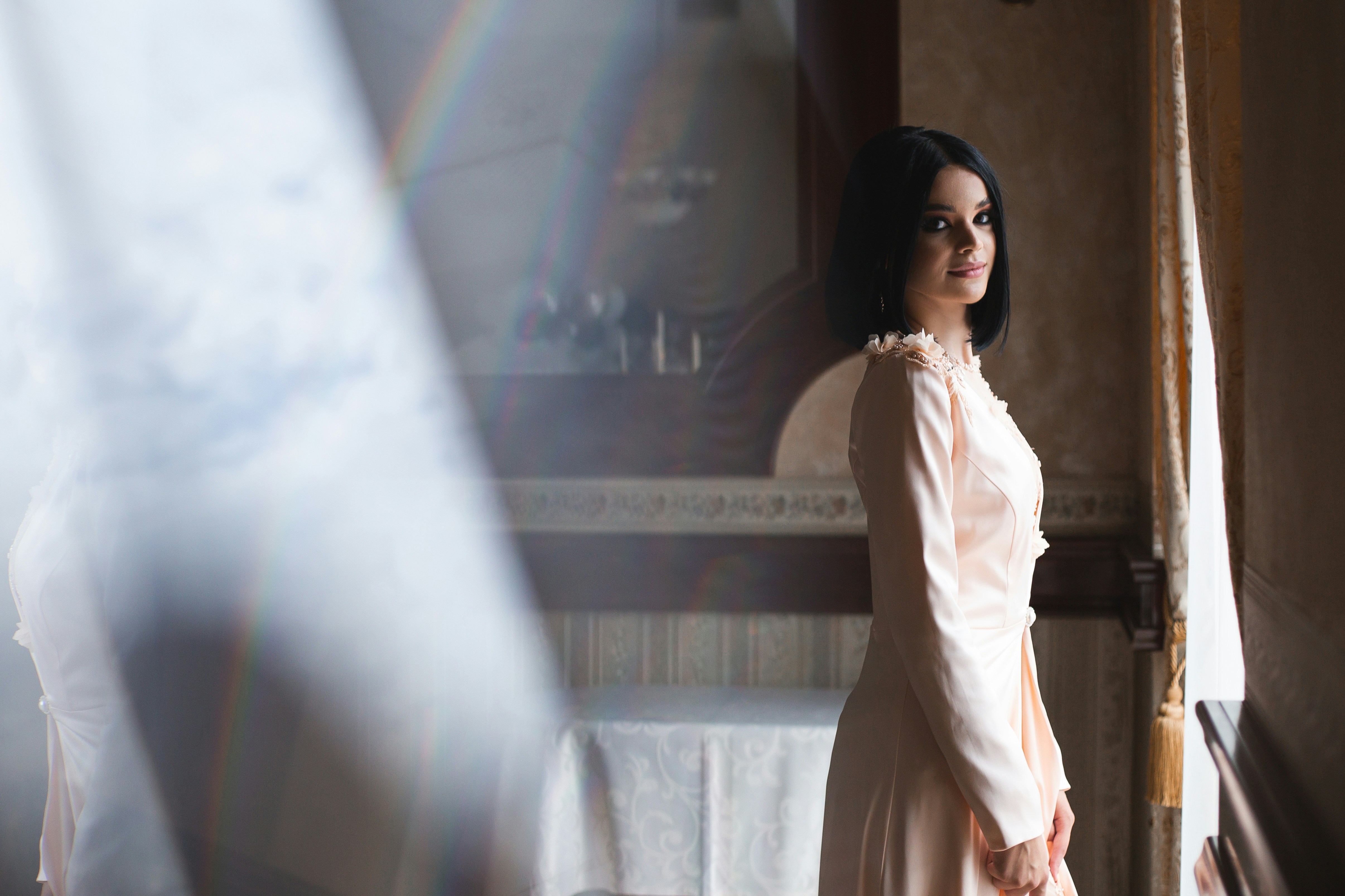 Girl by window, looking at camera. | Woman in light pink dress looking out window