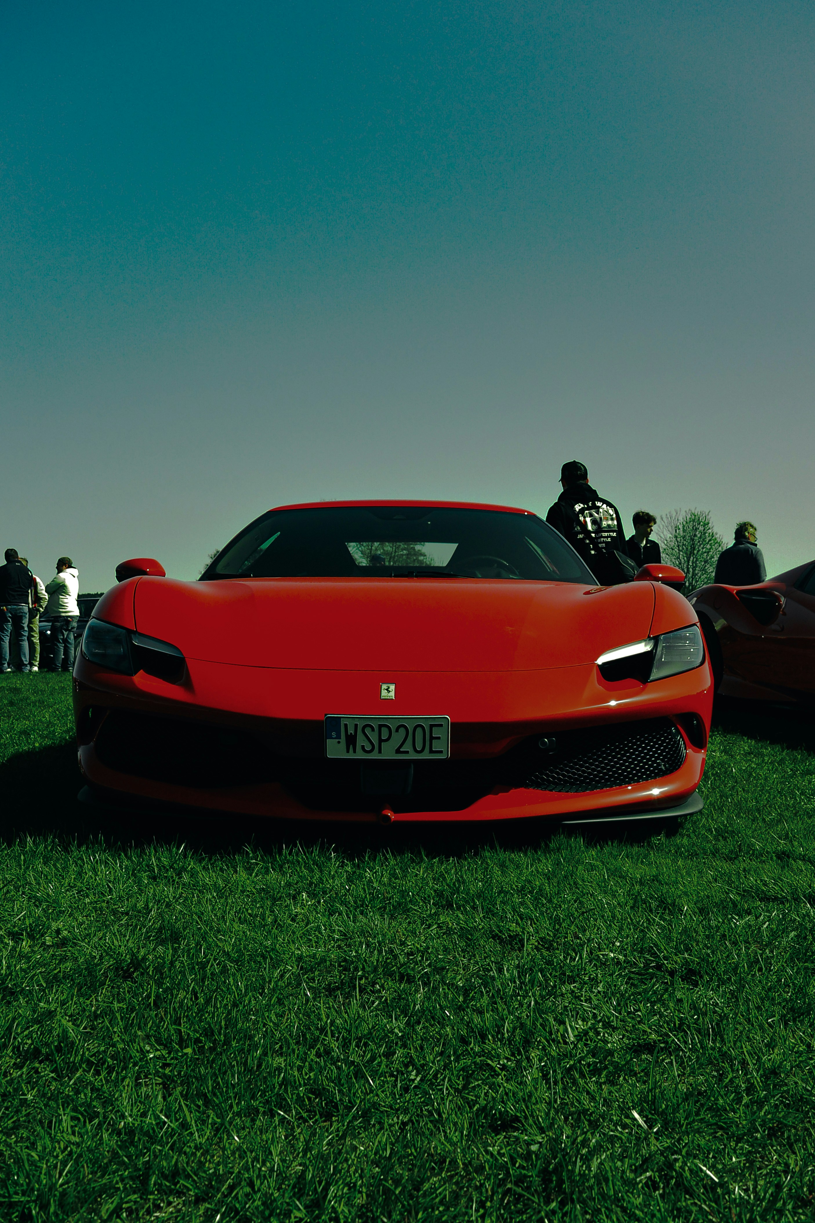 A striking red sports car showcased against a clear blue sky, surrounded by spectators on a grassy field.