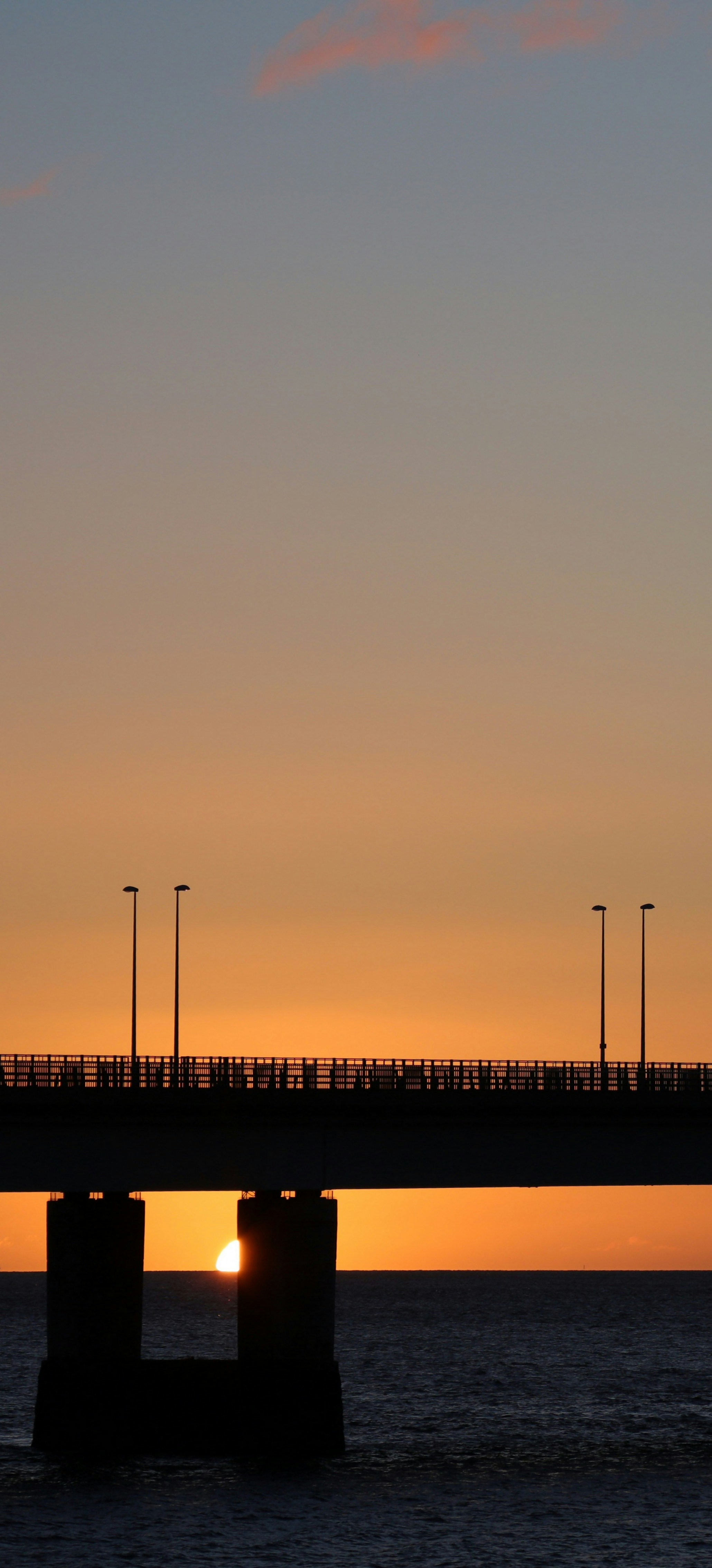Silhouette of a bridge against a vibrant sunset, with the sun partially visible below the structure, casting a serene glow on the water.