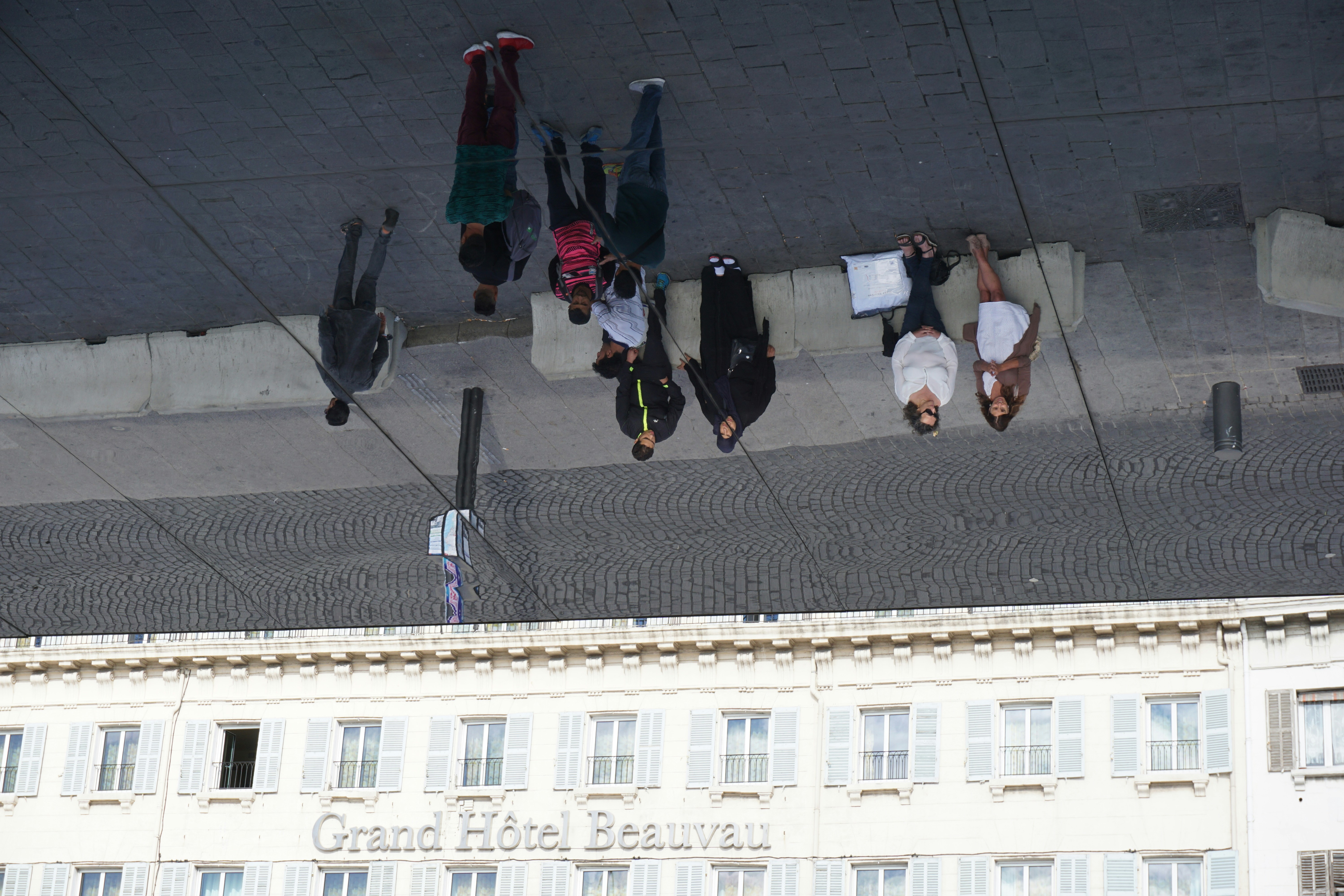 People walking reflected in a puddle beneath the Grand Hôtel Beauvau, showcasing urban life and architecture. The scene captures a moment of interaction between nature and city.