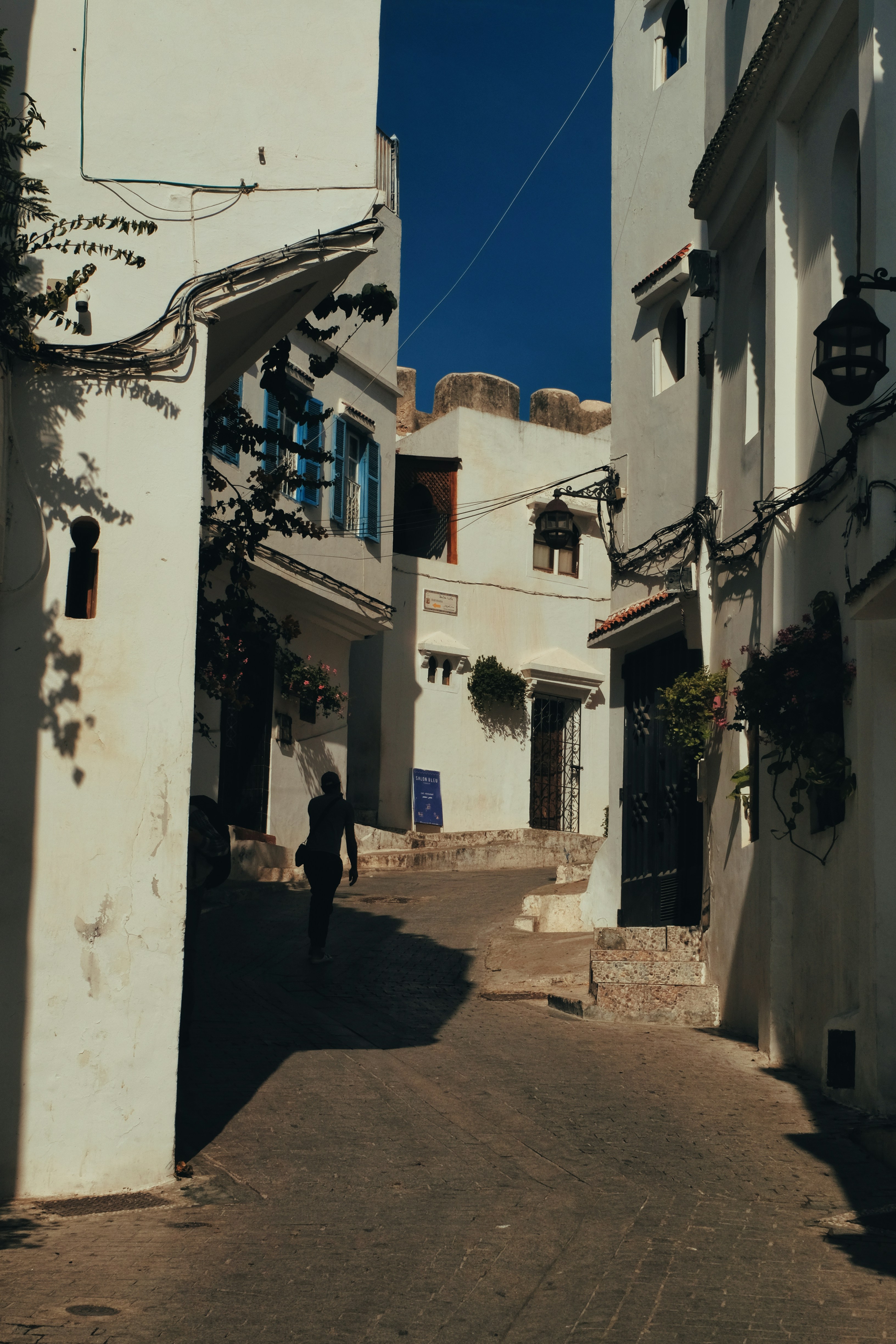 A lone figure walks down a narrow, sun-drenched street.