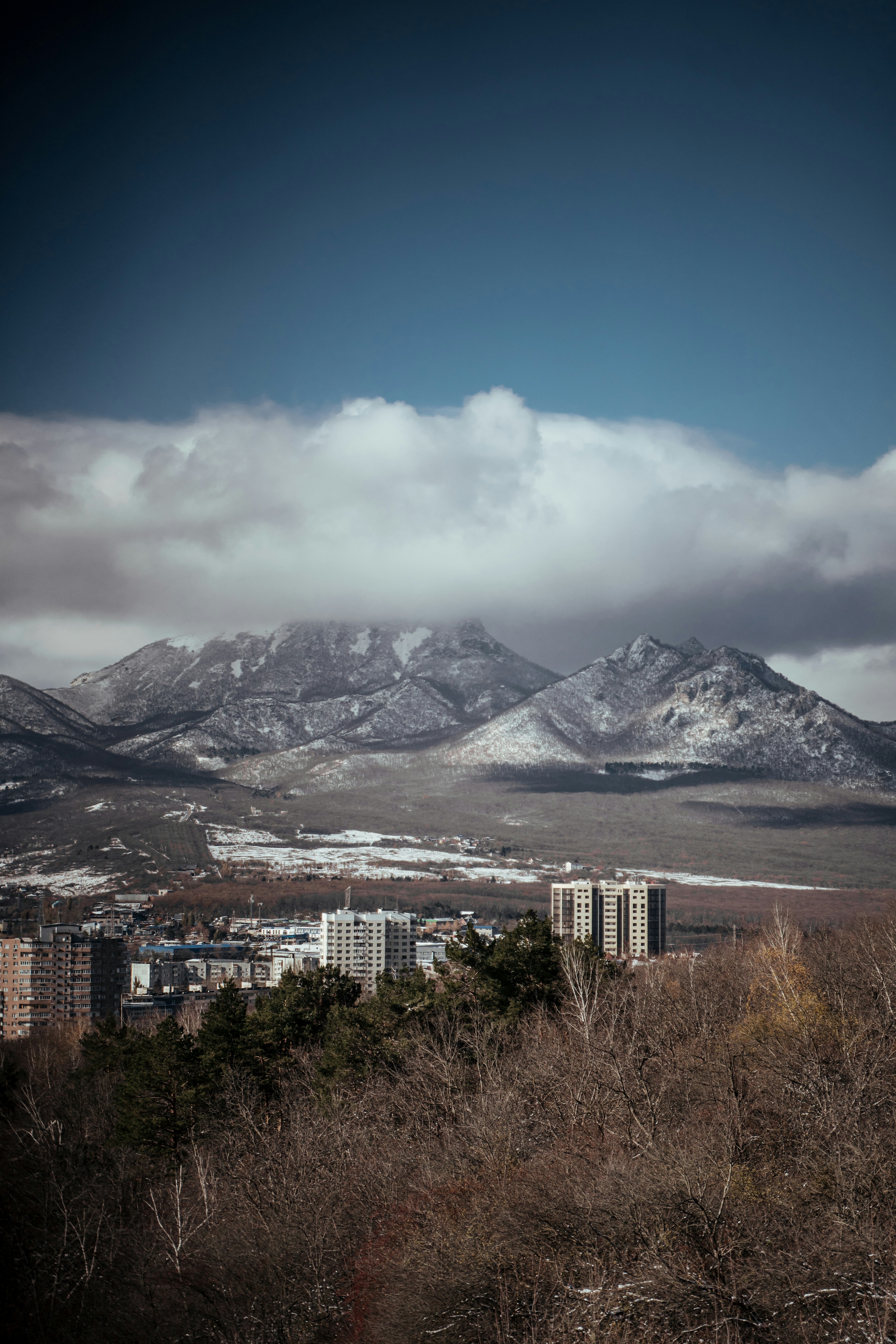 Snow-capped mountains loom over a city under cloudy skies.