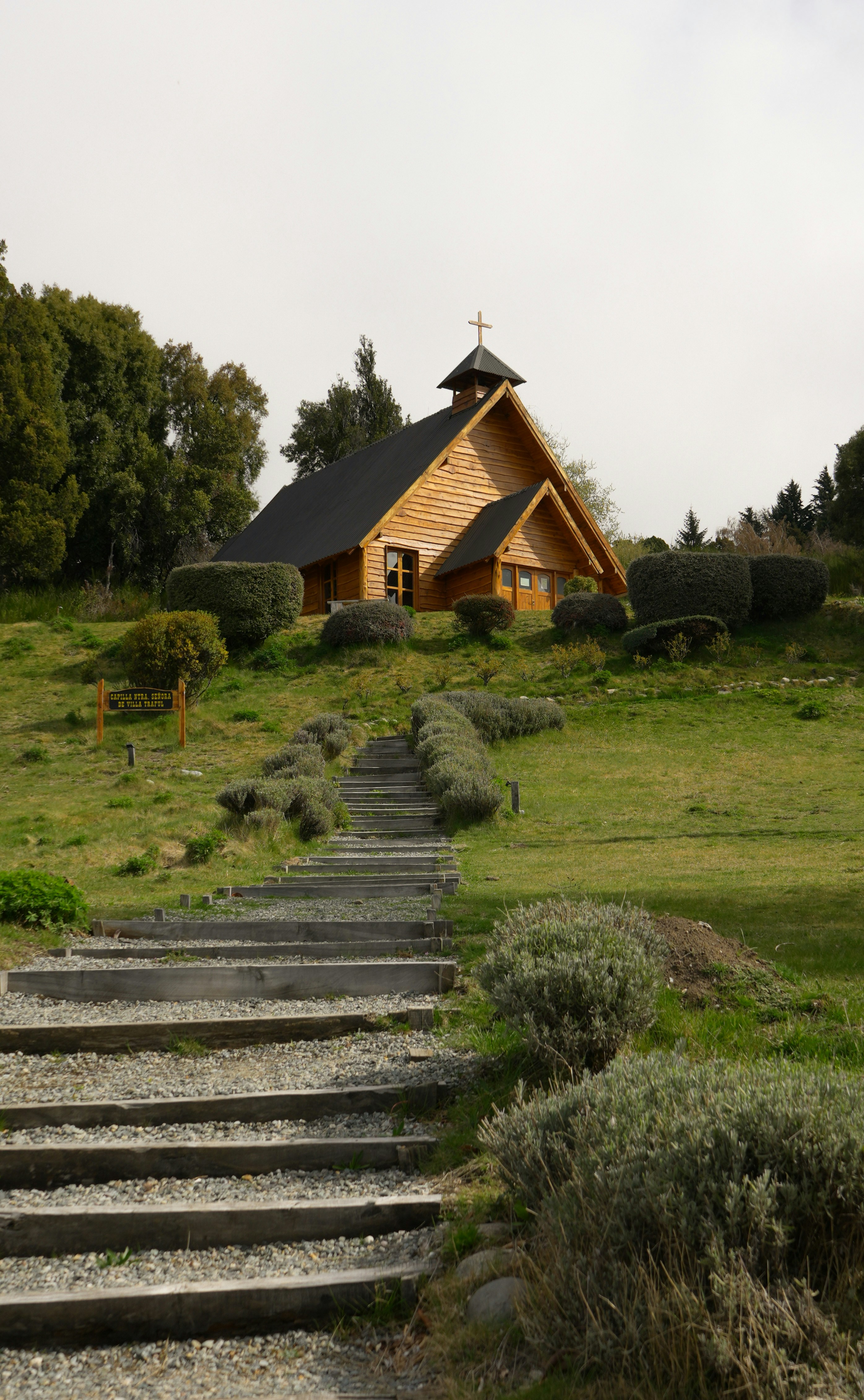 A charming wooden chapel nestled on a hillside, surrounded by manicured shrubs and a winding stone pathway leading up to it.