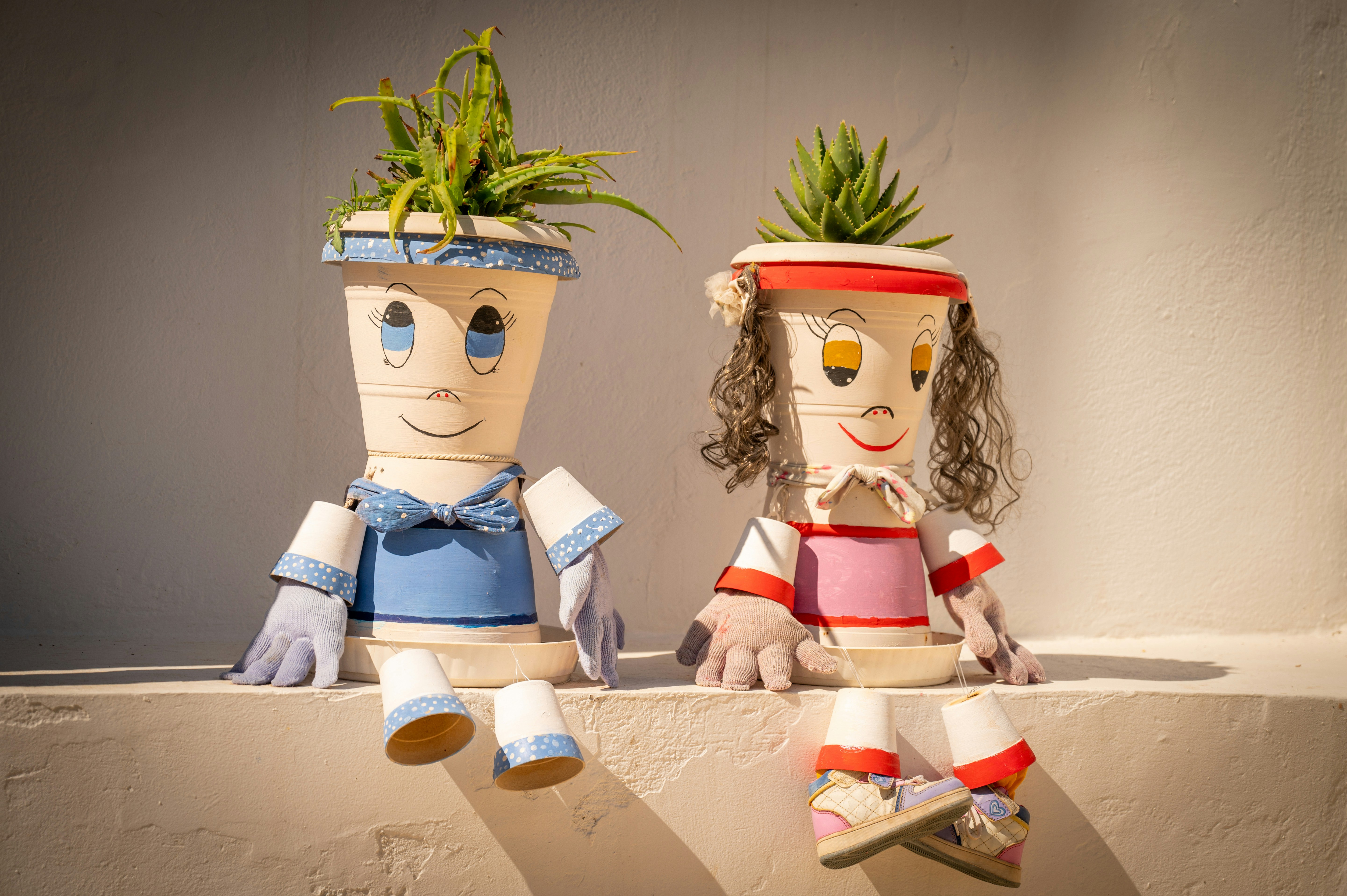 Two playful planters with faces and colorful outfits, adorned with small plants, sit side by side on a ledge.