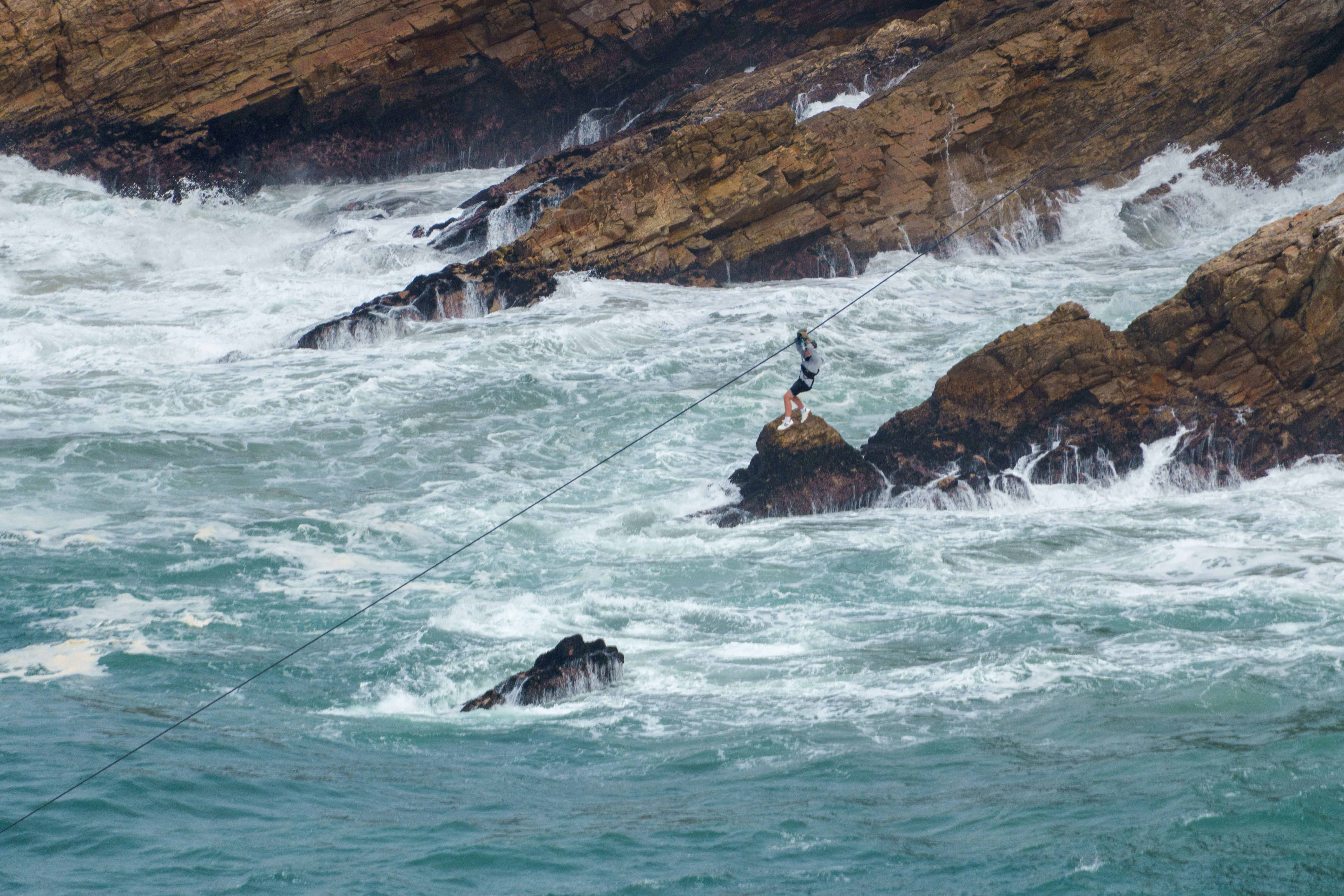 A person precariously balancing on a rock amidst crashing waves, showcasing the raw power of nature. The scene captures the tension between human determination and the ocean's might.