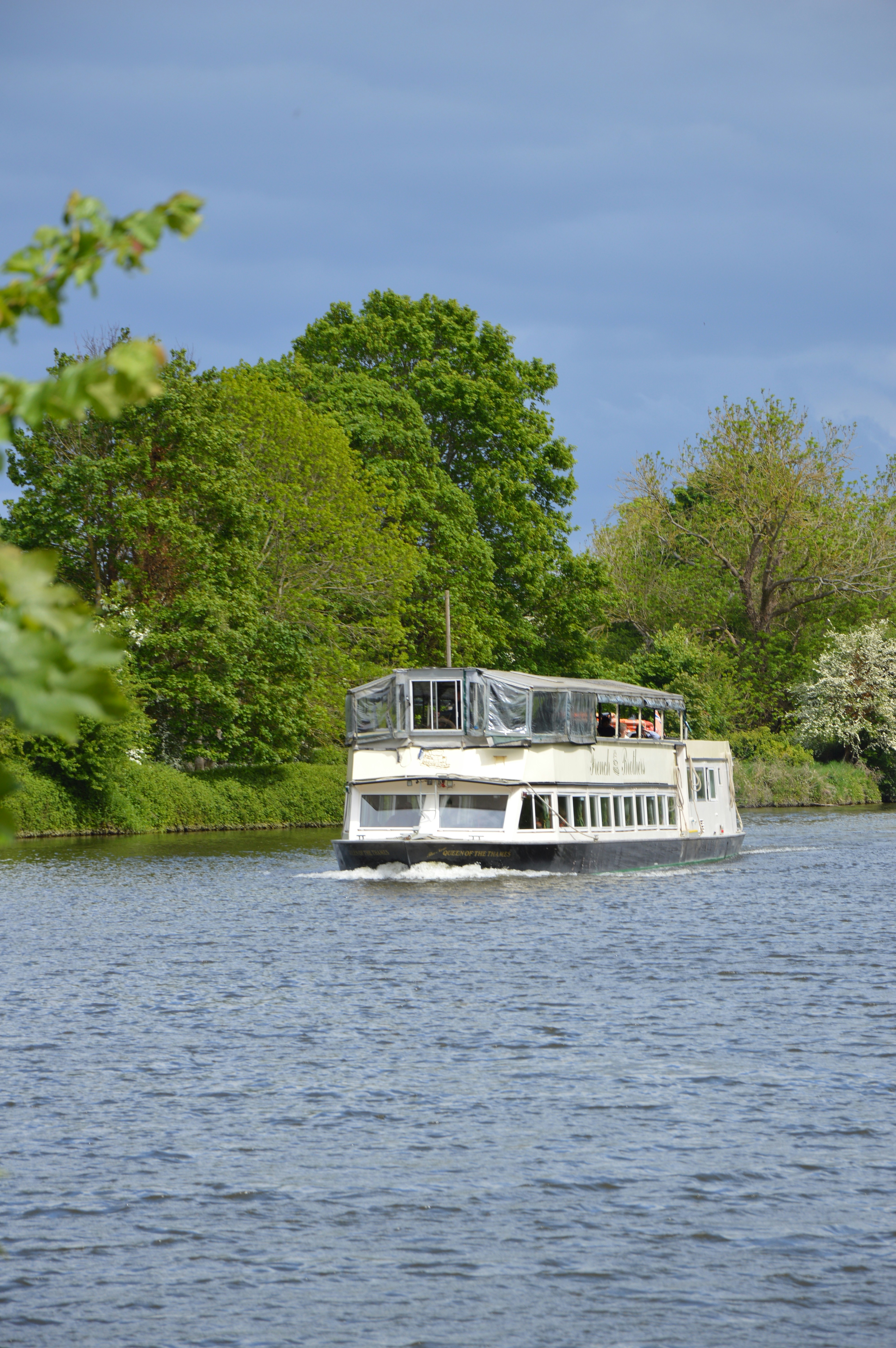A white boat glides smoothly across a tranquil river, surrounded by lush greenery and a cloudy sky.