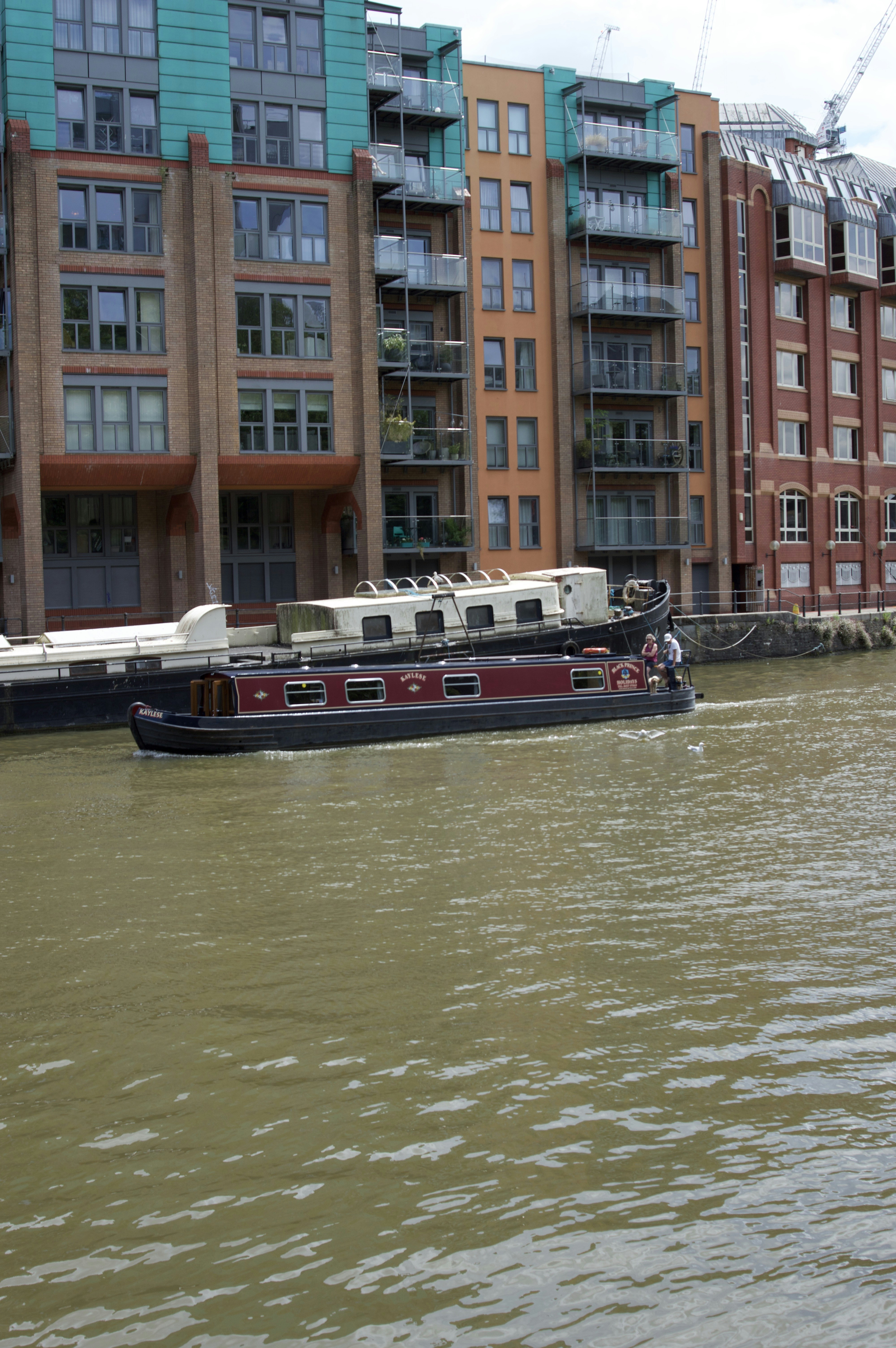 Narrowboat gliding through a tranquil canal bordered by modern residential buildings. Sunlight dances on the water's surface.