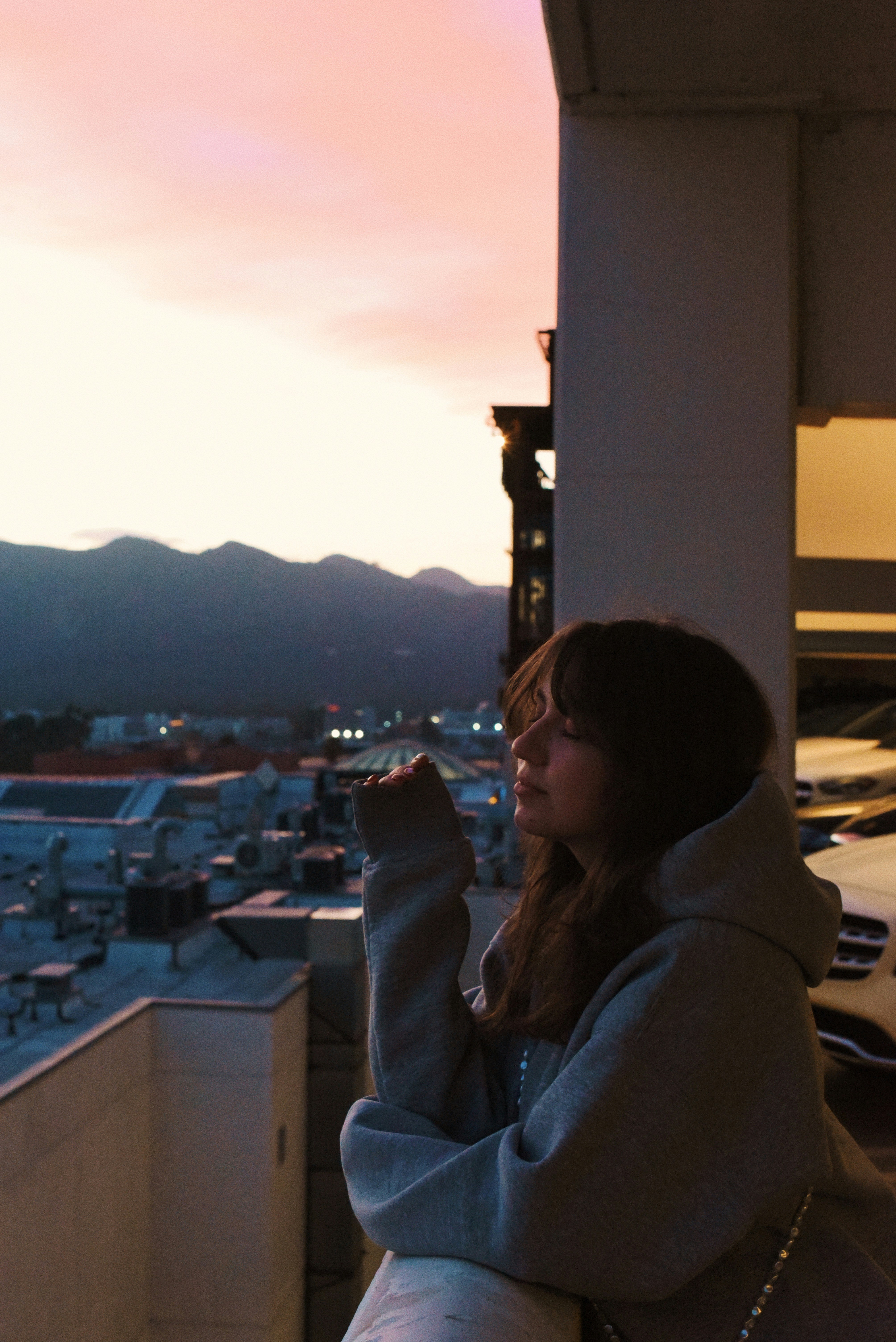 A contemplative figure in a cozy hoodie gazes into the horizon as twilight descends, with mountains silhouetted against a colorful sky.