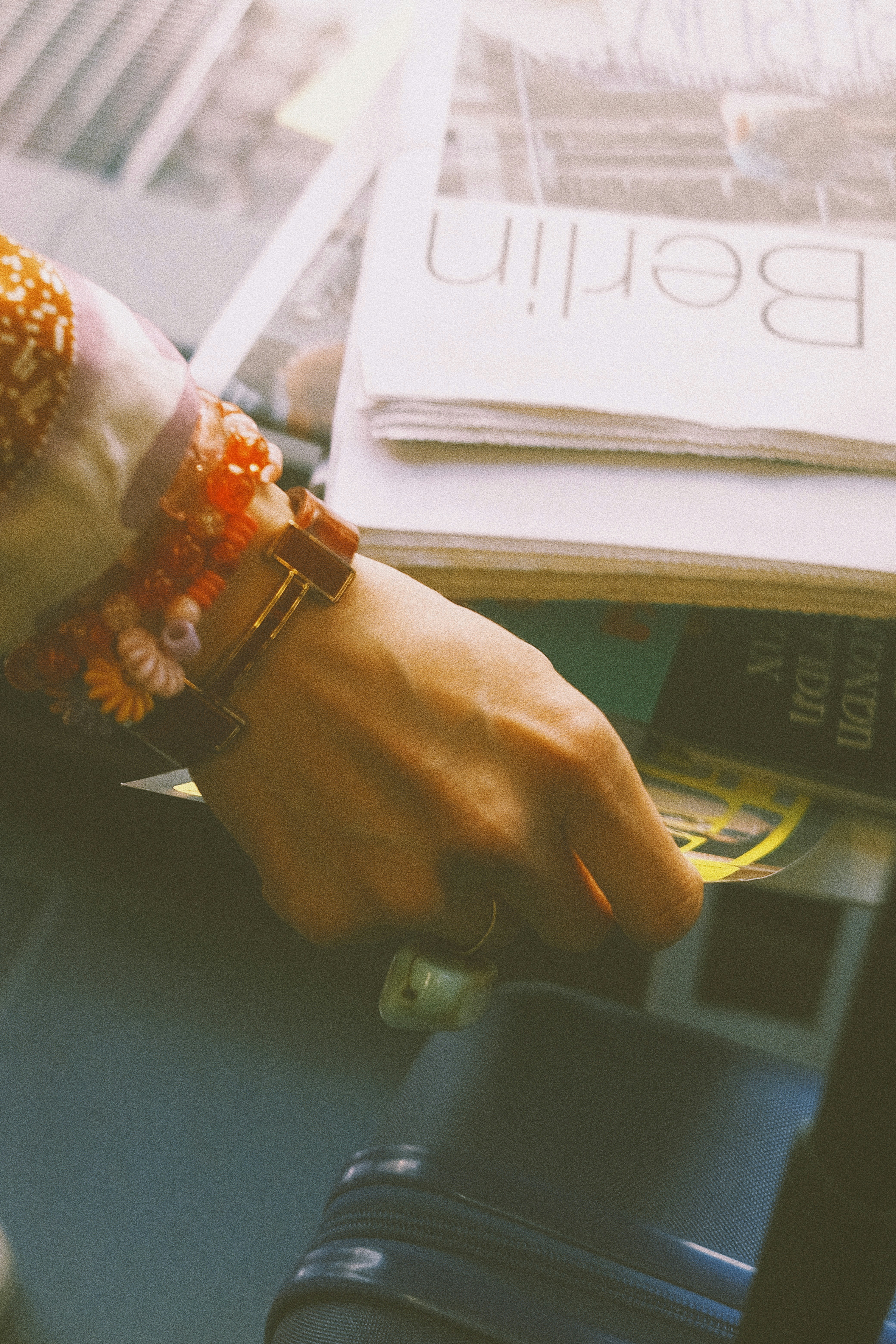 Close-up of a hand adorned with colorful bracelets, resting on a stack of magazines and a suitcase. The scene captures a fleeting moment of everyday life.