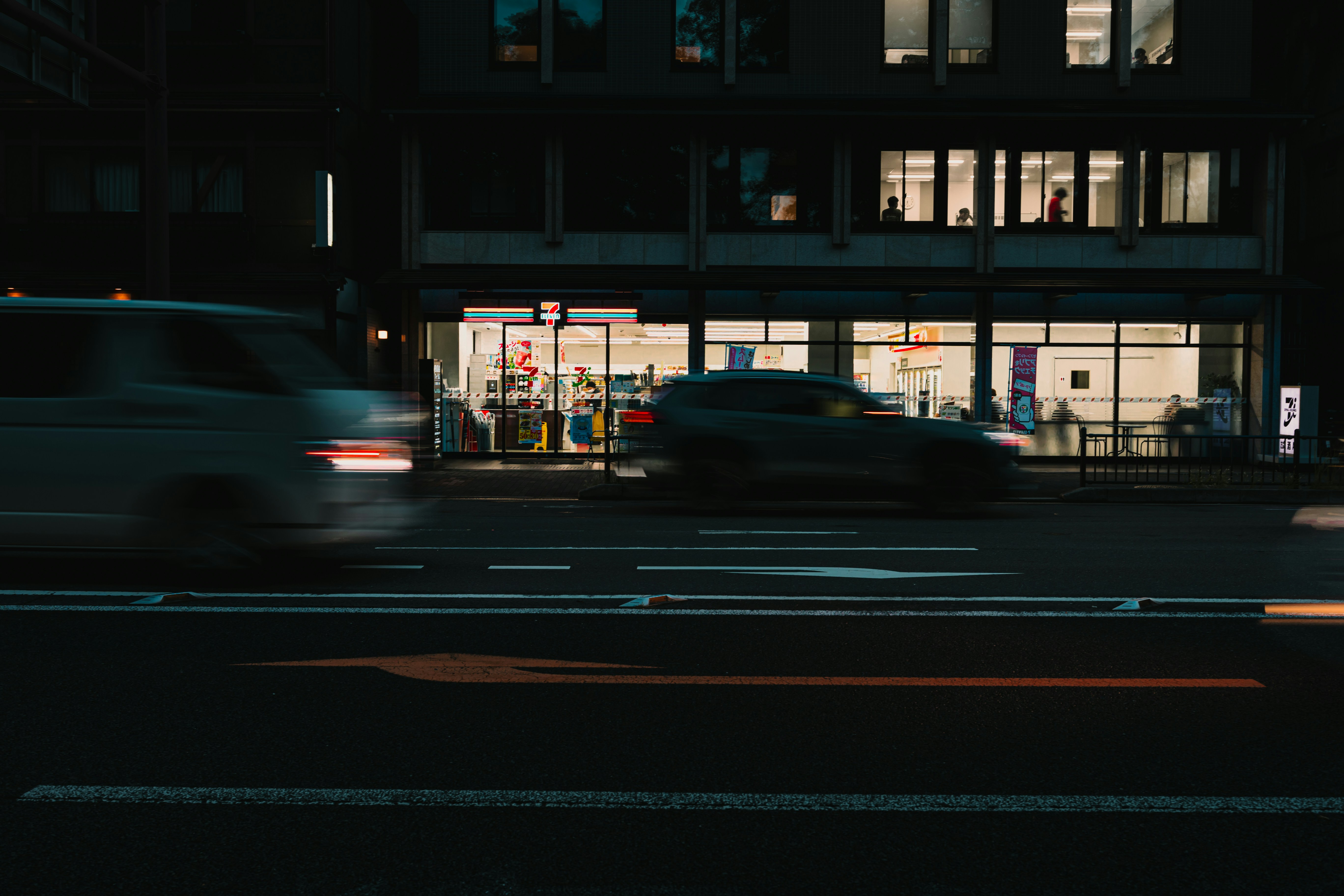 Blurred vehicles pass by a brightly lit storefront, showcasing the contrast between movement and stillness in an urban setting.