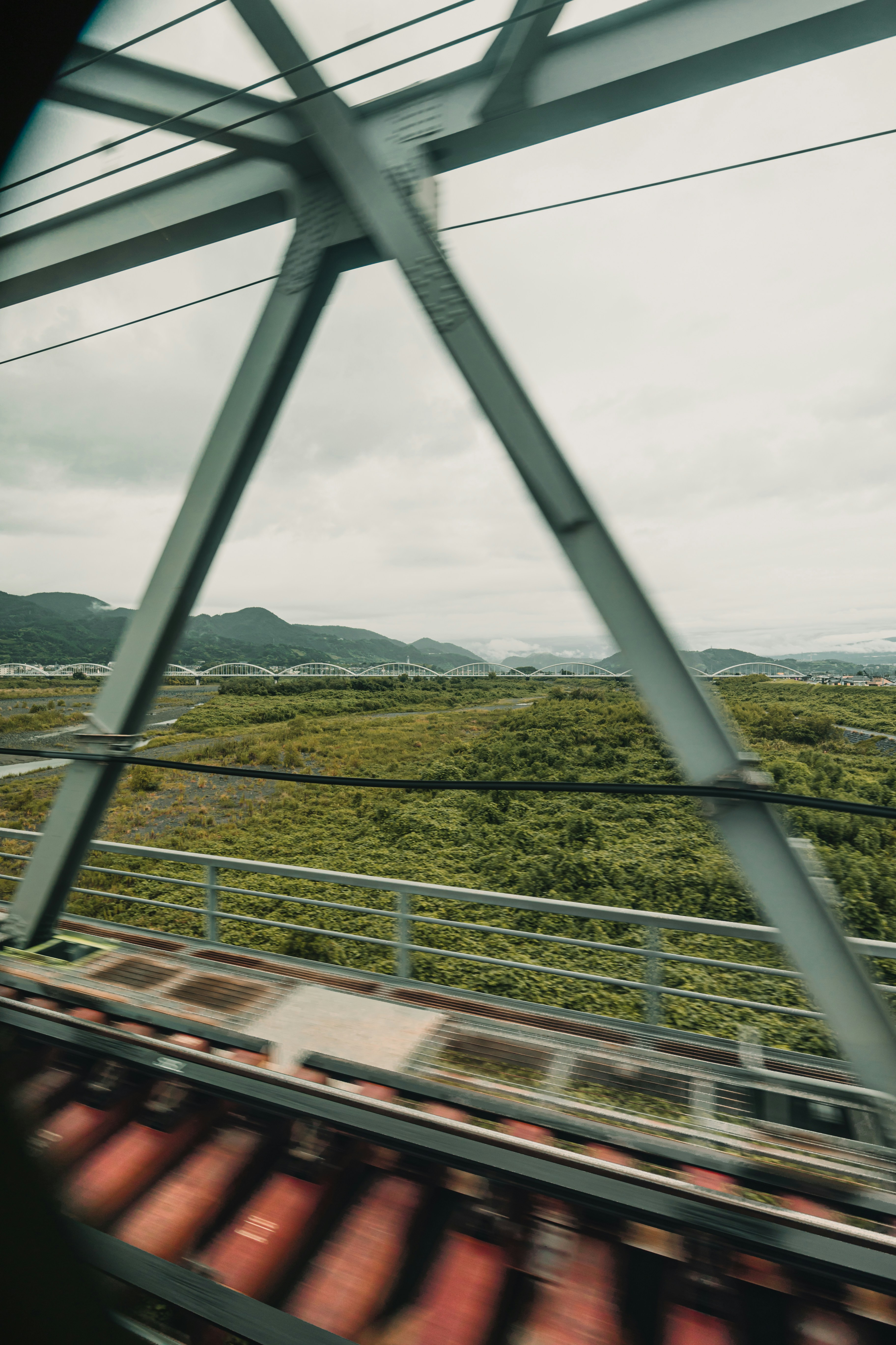 View from a moving train showcasing a lush landscape framed by a metallic bridge structure. The scene captures the interplay between nature and human engineering.
