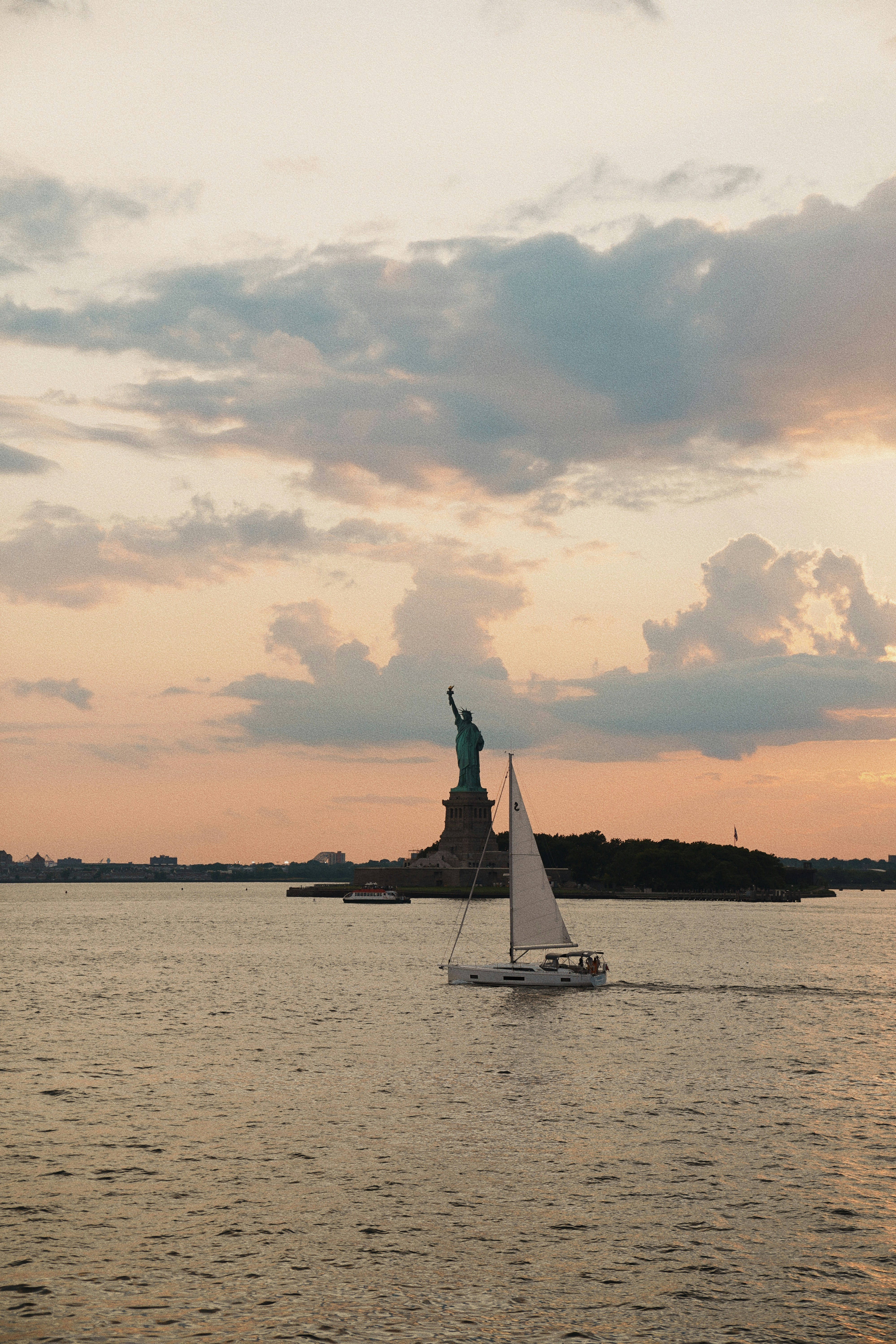 Dusk Over Liberty: A Sailboat Crossing the Quiet Harbor