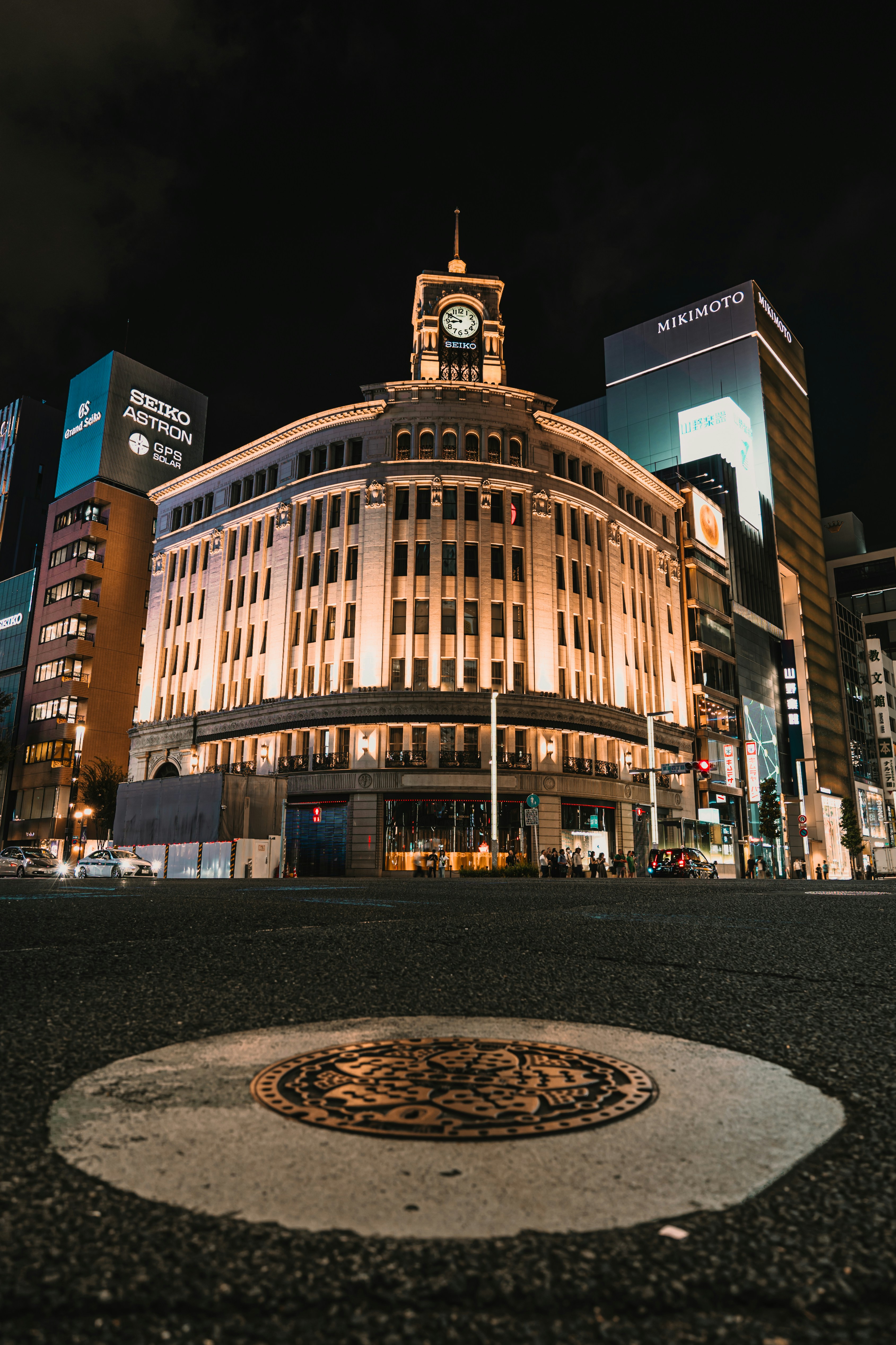 Nighttime photograph of a curved, illuminated building crowned by a clock tower, framed by tall billboards in a busy city street. The foreground features a textured manhole cover, anchoring the urban scene.