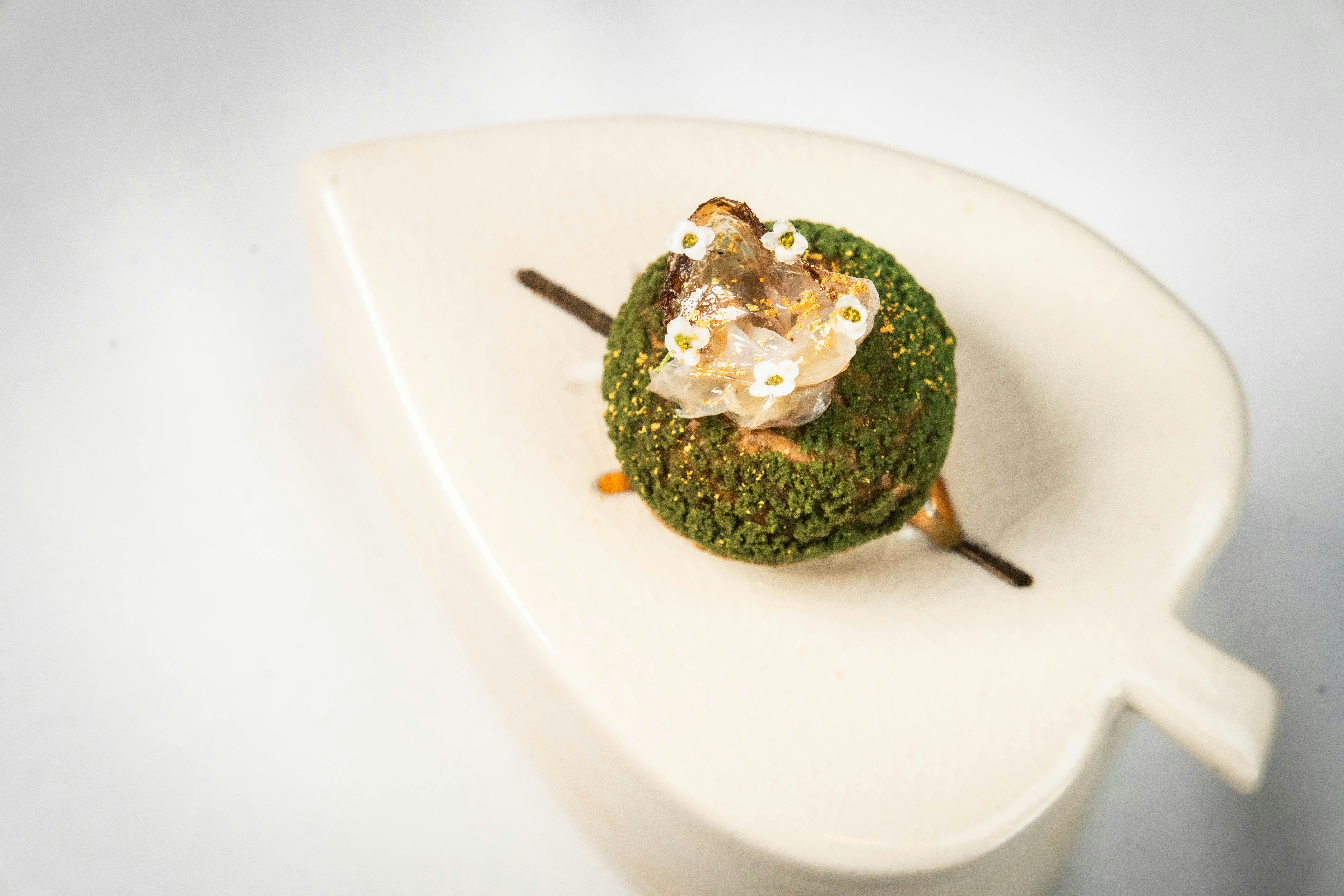 Artfully presented gourmet dish featuring a vibrant green base topped with delicate flowers and a golden accent. Served on a uniquely shaped plate.