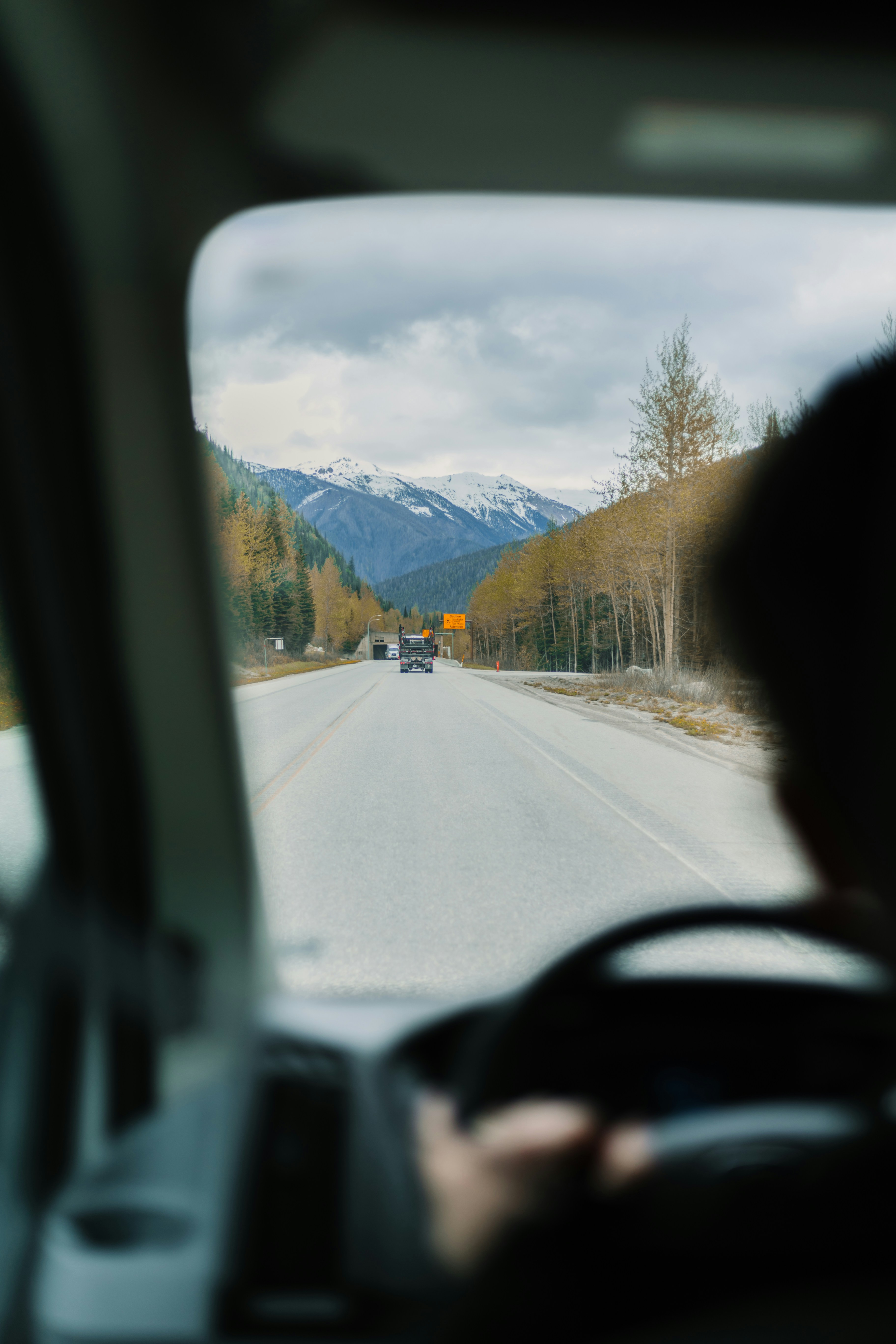 Vehicle navigating a winding road framed by snow-capped mountains and autumn trees, hinting at a scenic adventure ahead.