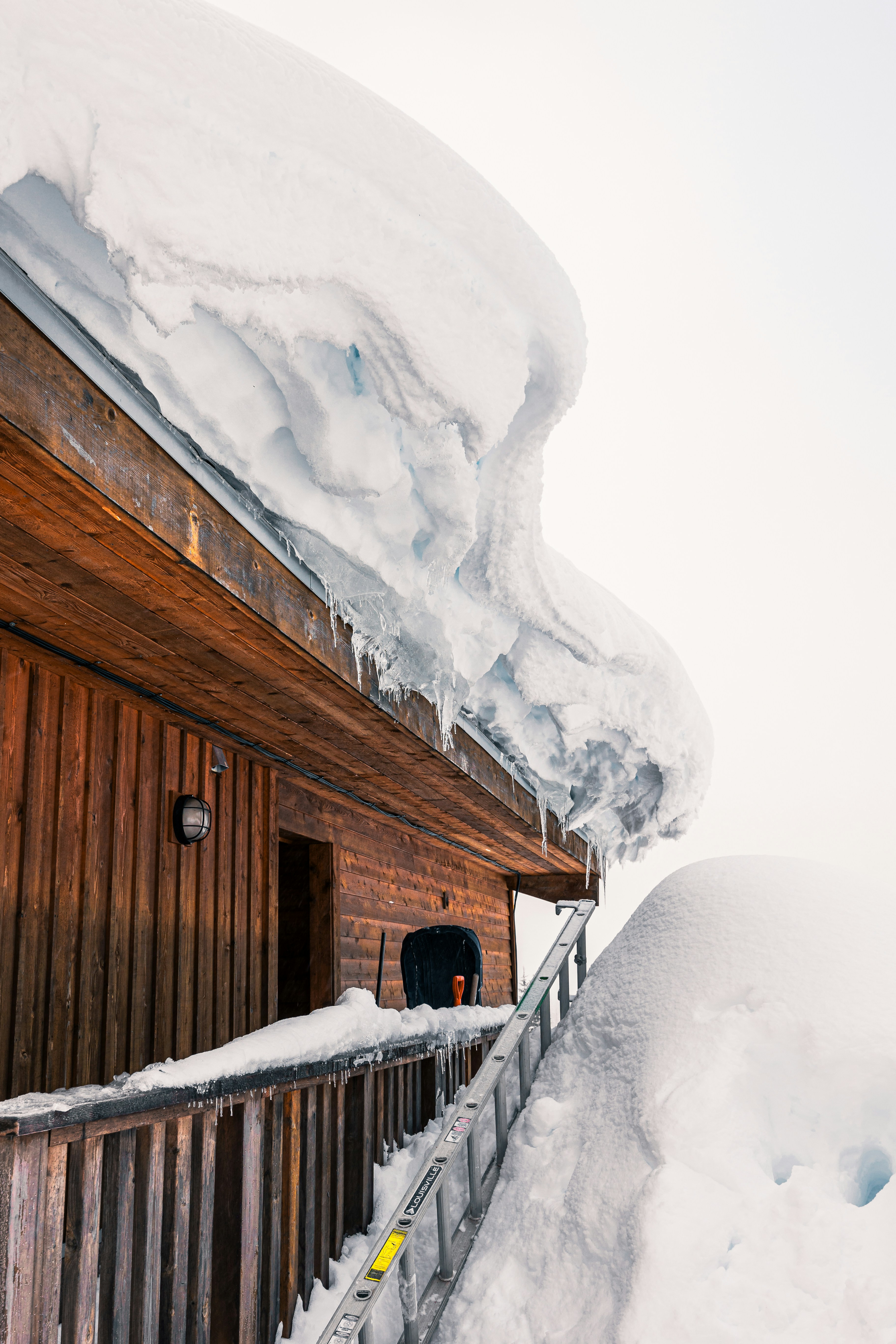 Extreme snow cornice overhangs the roof and railing of a wooden cabin, with an aluminum ladder visible on the railing. | Wooden cabin with heavy snow accumulation on roof