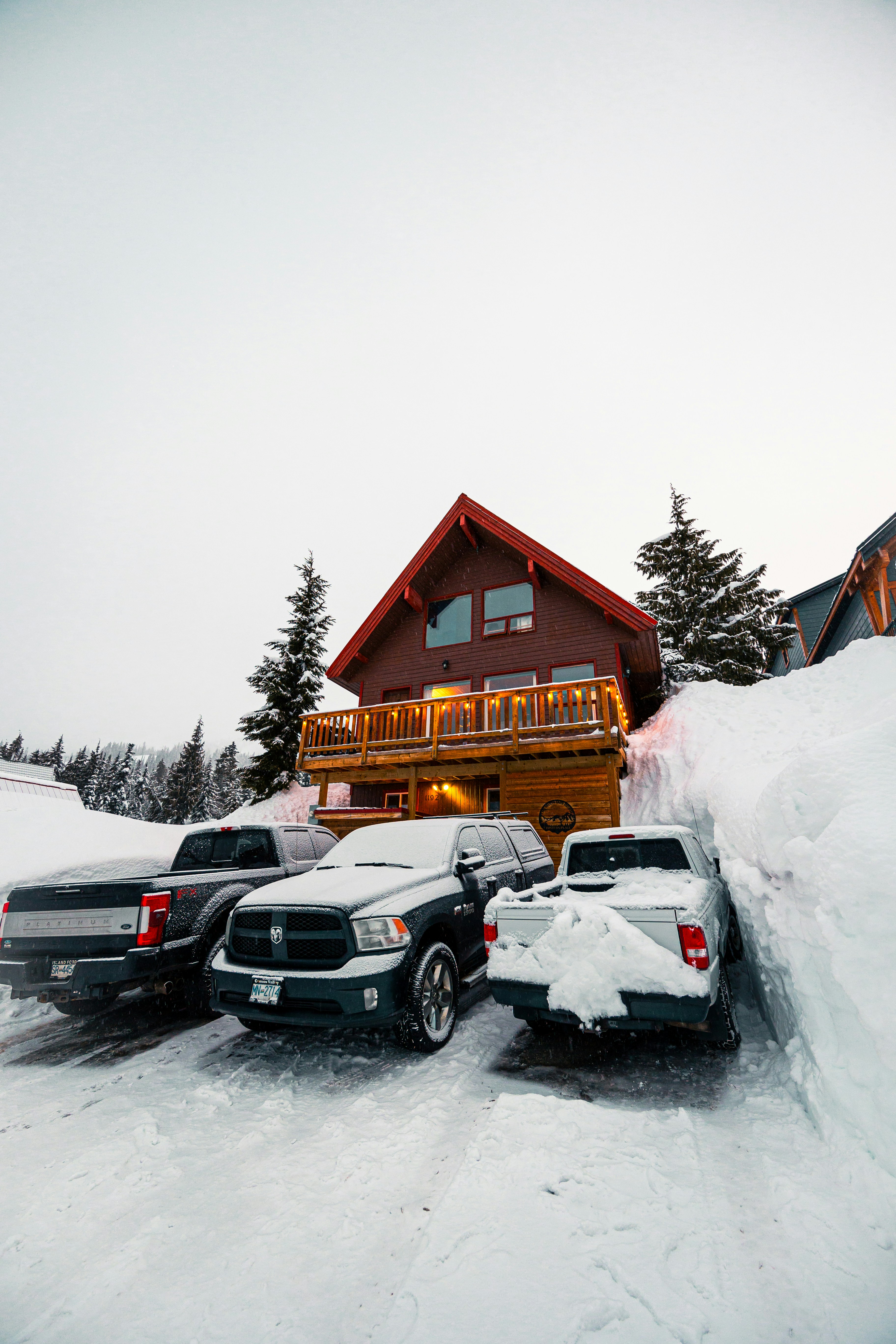 A wooden cabin with a red roof and balcony, framed by deep snowbanks and three parked pickup trucks.