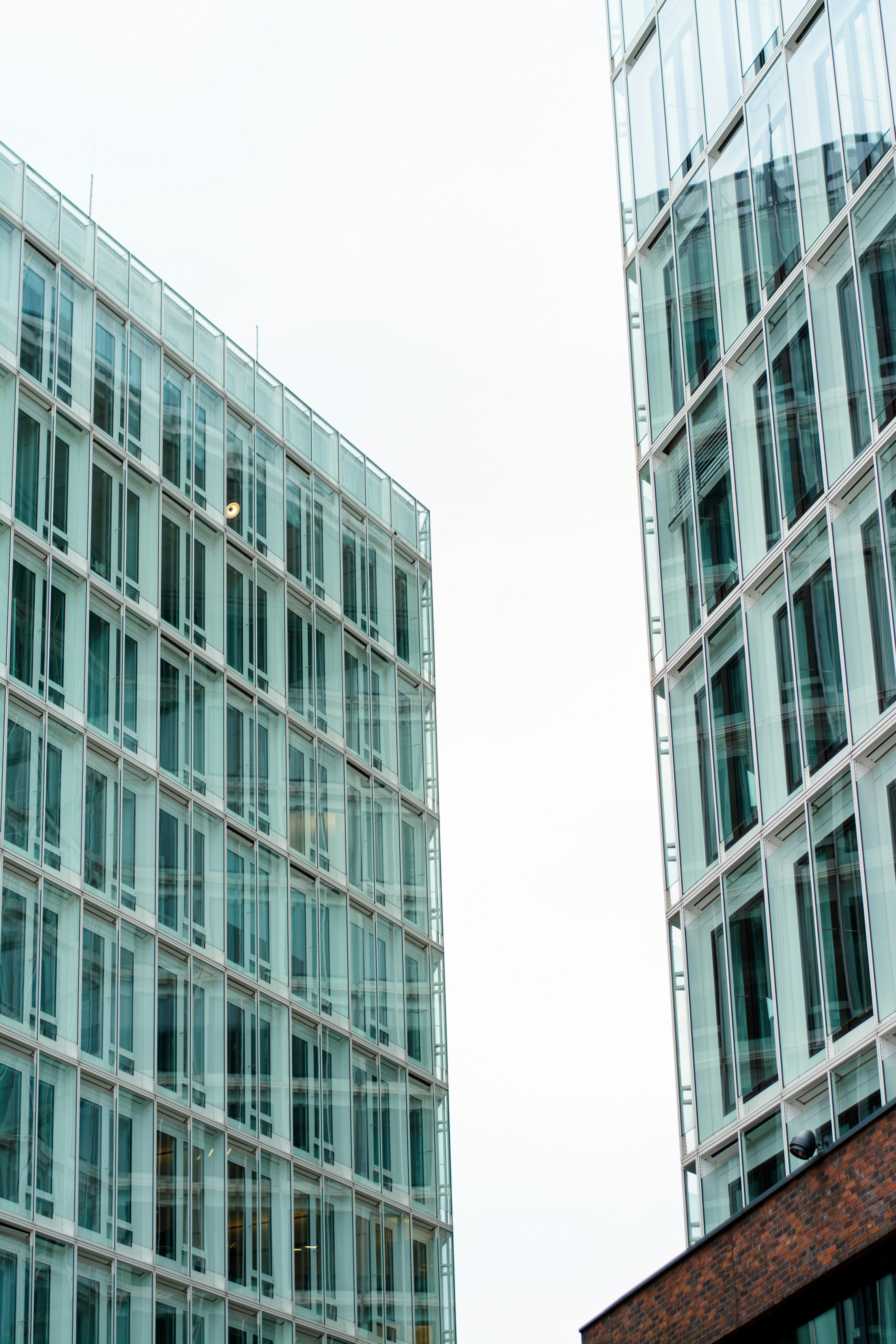 Two contemporary glass buildings converge towards the sky, showcasing their reflective façades against a soft gray backdrop.
