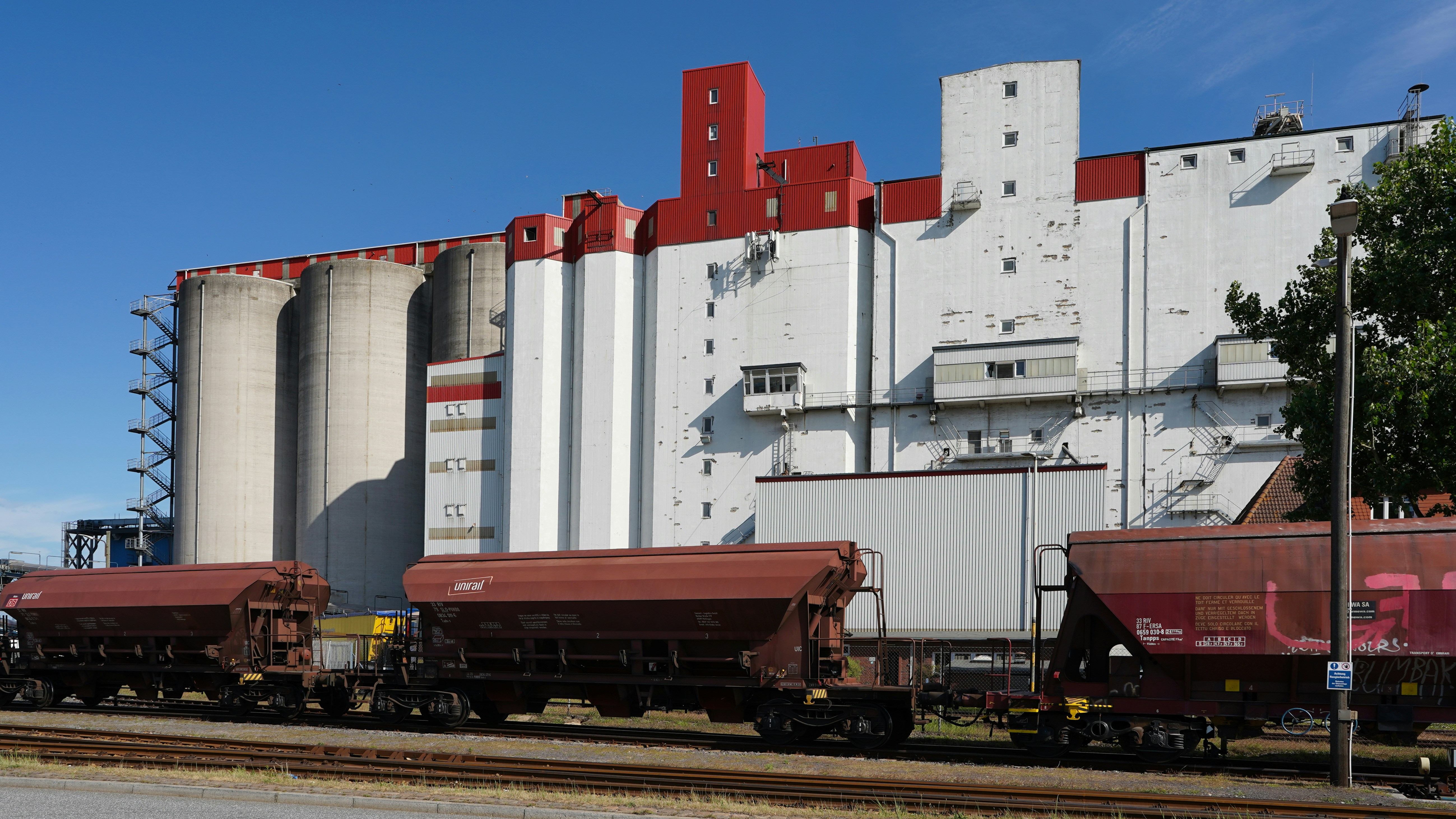 Weathered grain silos stand tall against a clear blue sky, with freight cars parked in the foreground. The scene captures the essence of industrial life and transportation.