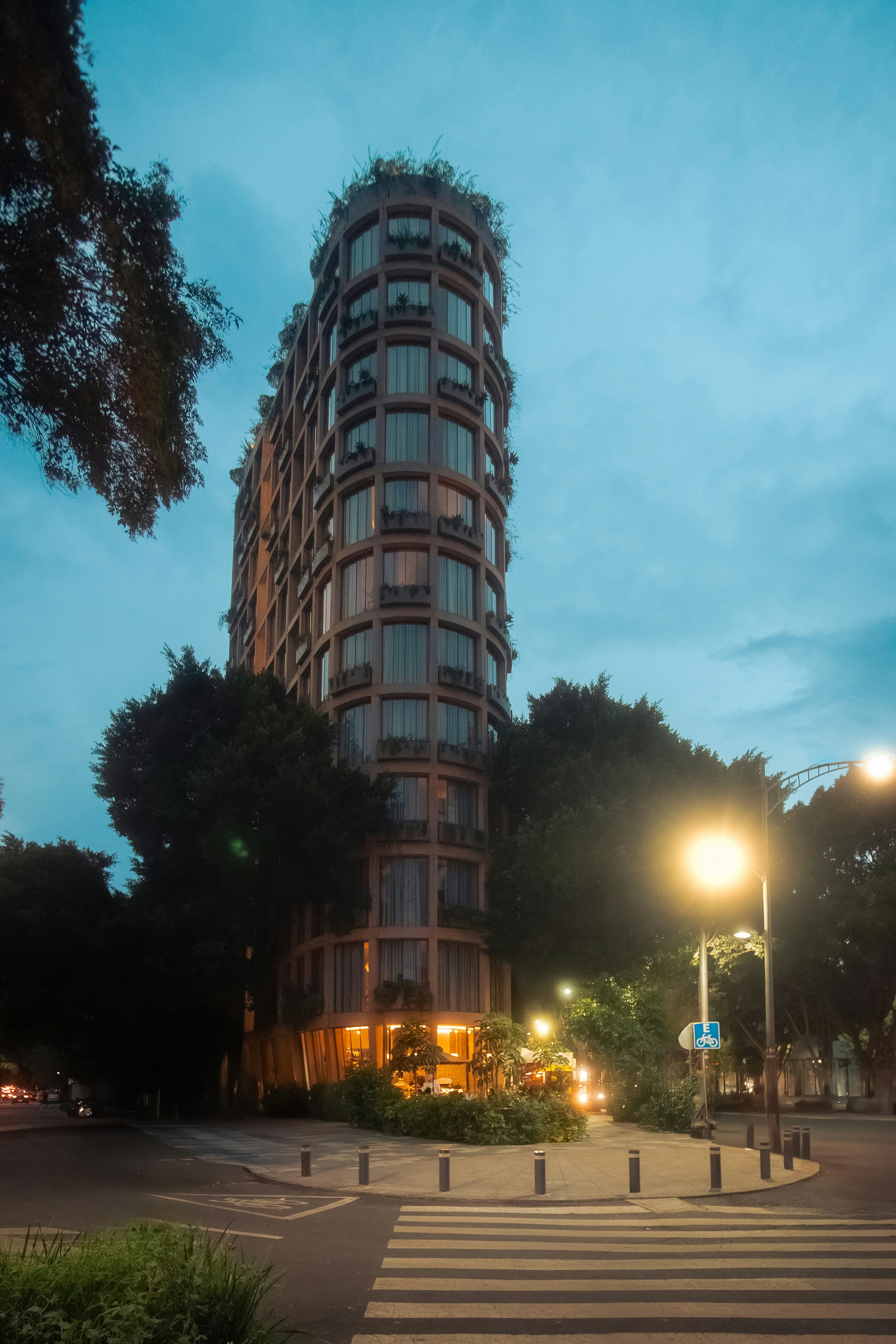 A modern cylindrical building surrounded by lush greenery and streetlights, showcasing the blend of architecture and nature in an urban setting.