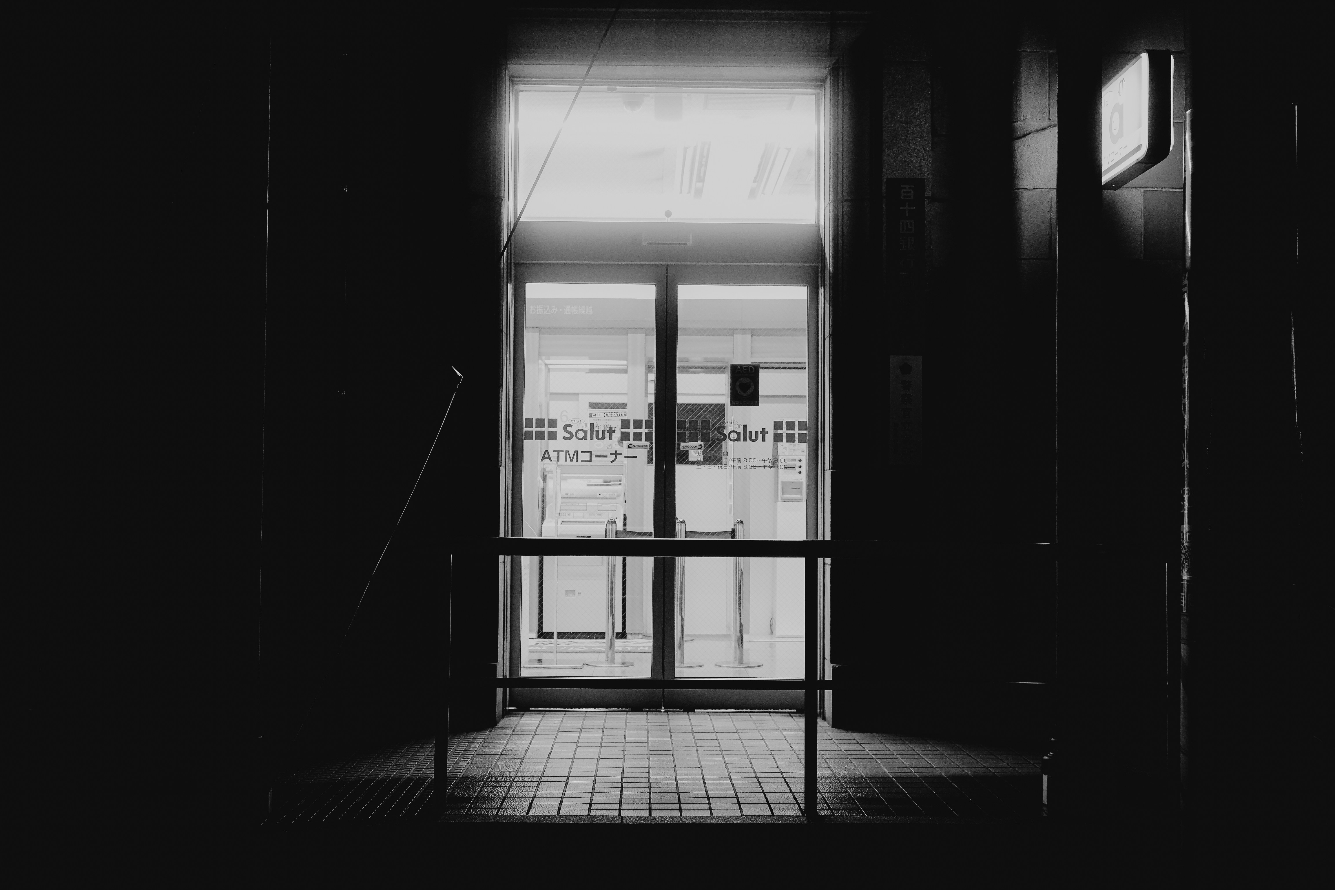 Dimly lit entrance to an ATM area, framed by dark walls and a stark contrast of light. The scene evokes a sense of solitude and anticipation.