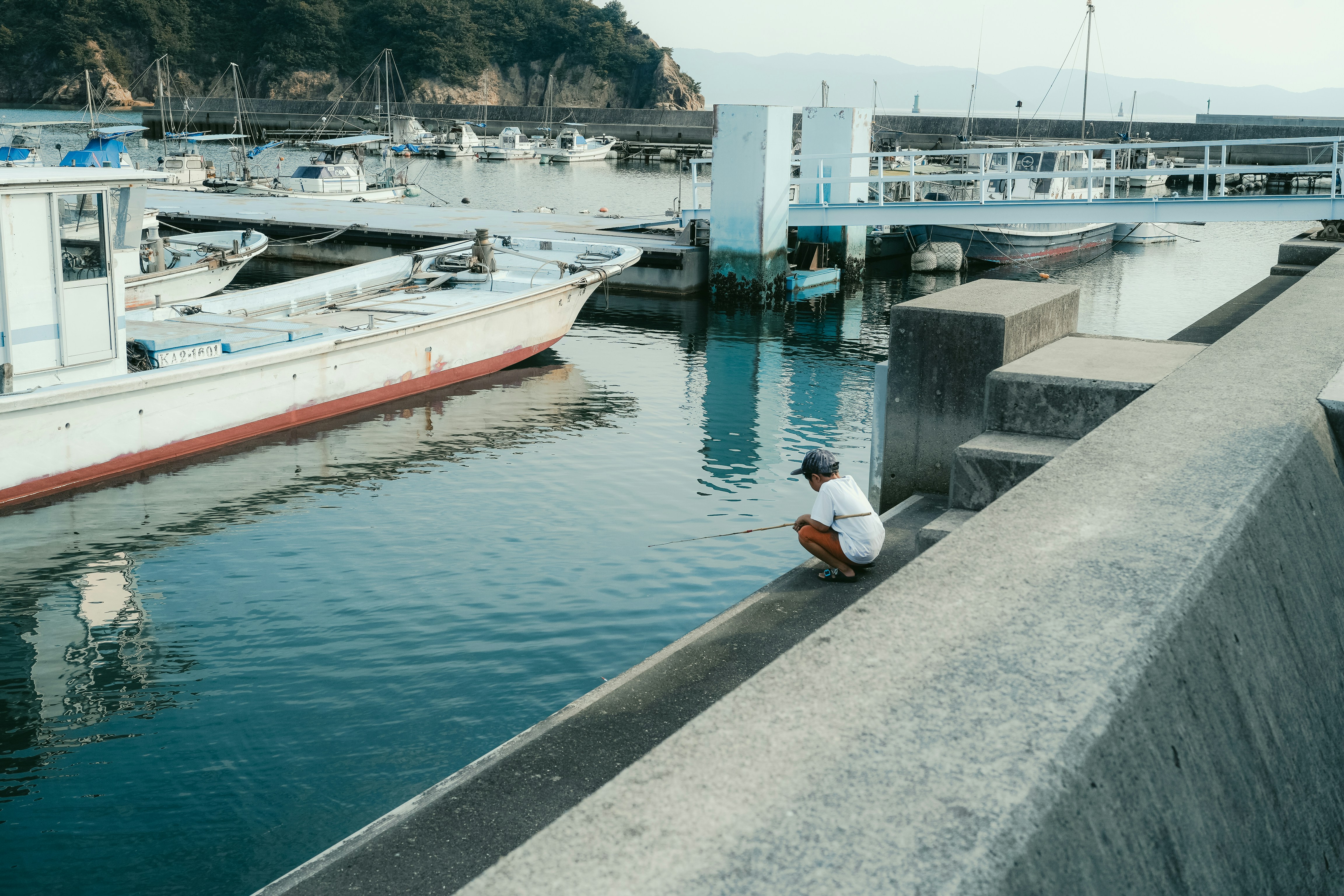 Man fishing by the harbor with boats and buildings.