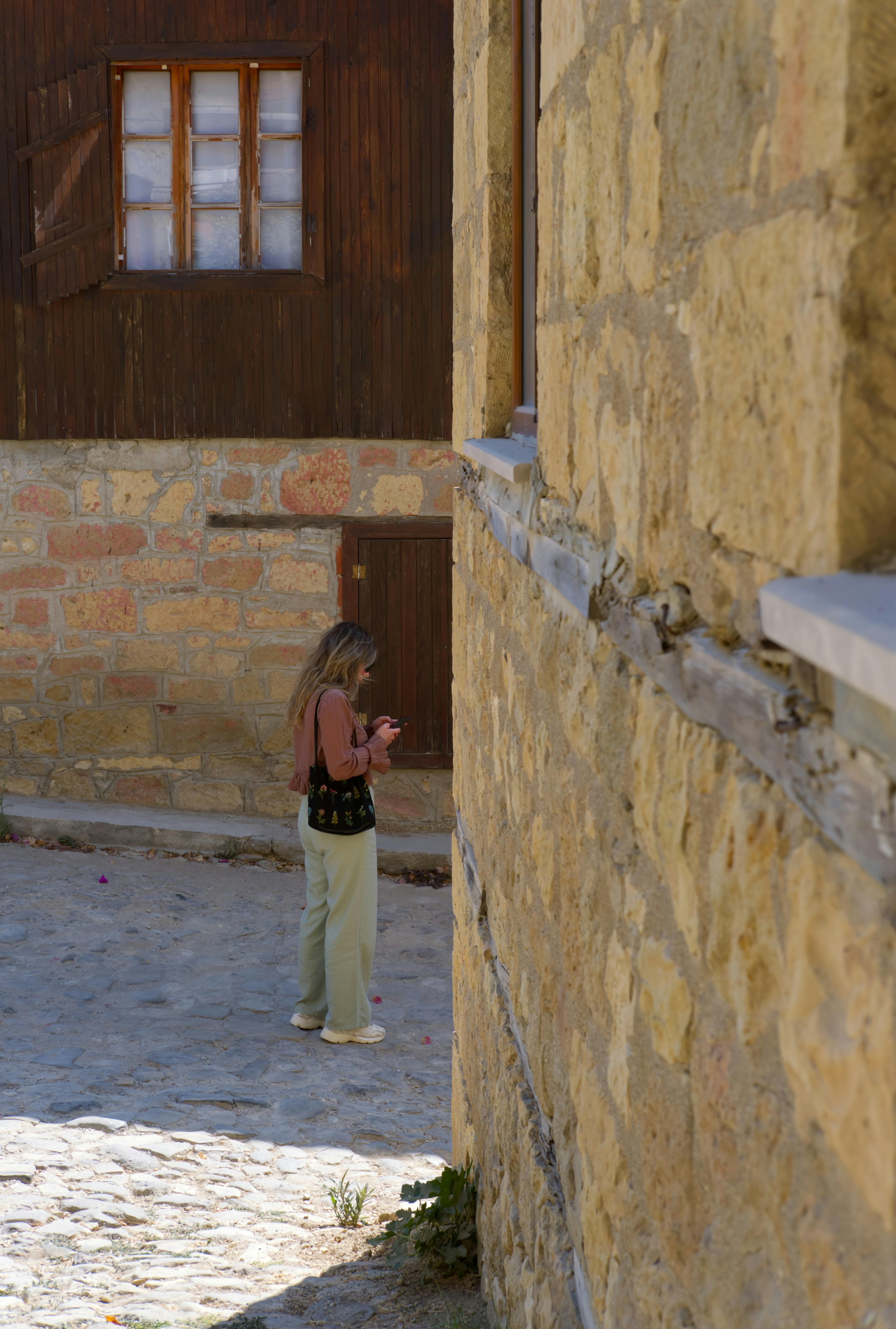 A young woman stands absorbed in her phone, framed by rustic stone walls in a quiet alley. The sunlight casts soft shadows, enhancing the tranquil atmosphere.