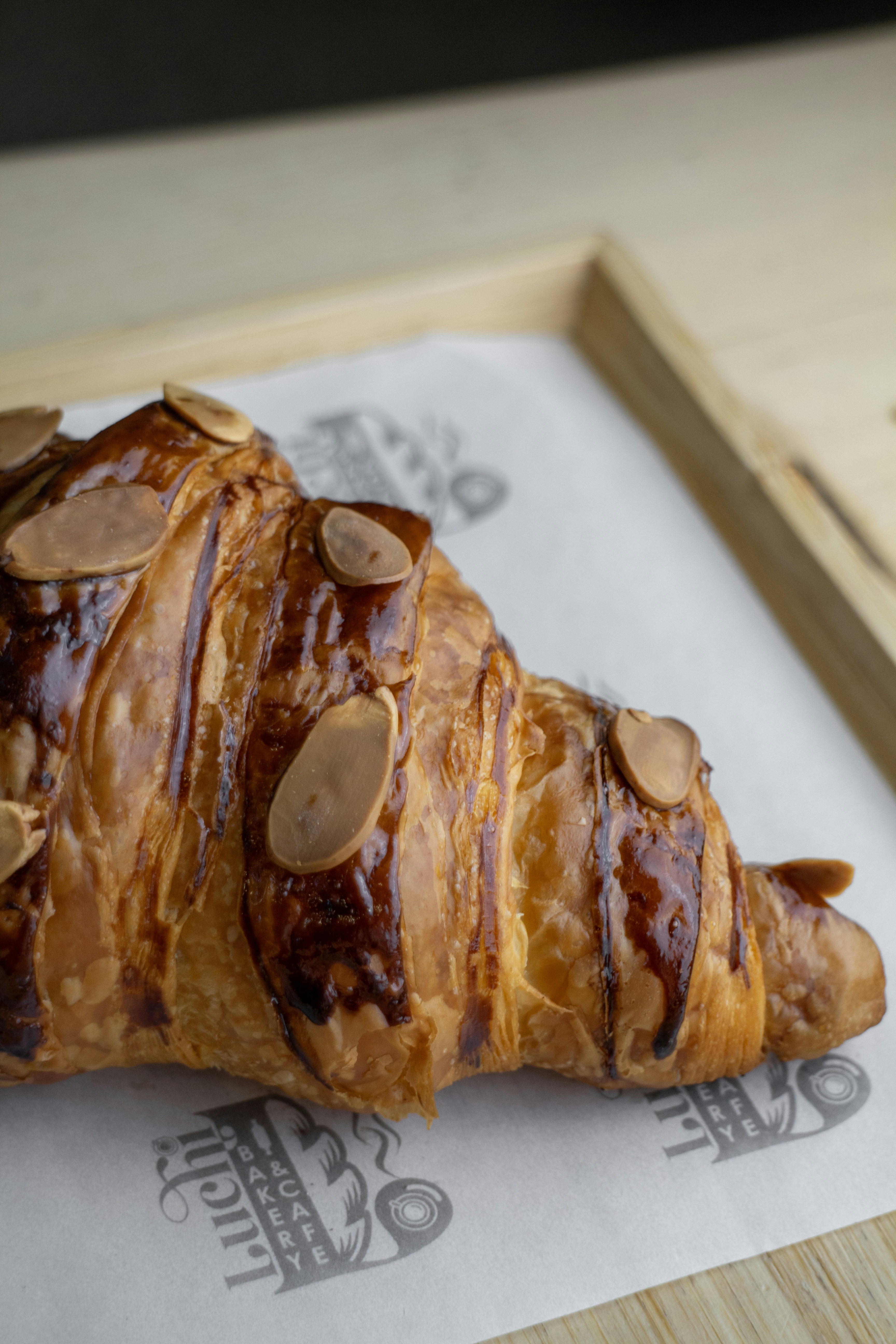 Freshly Baked Croissant with Buttery Layers – Morning Light | Almond croissant on paper with wooden tray