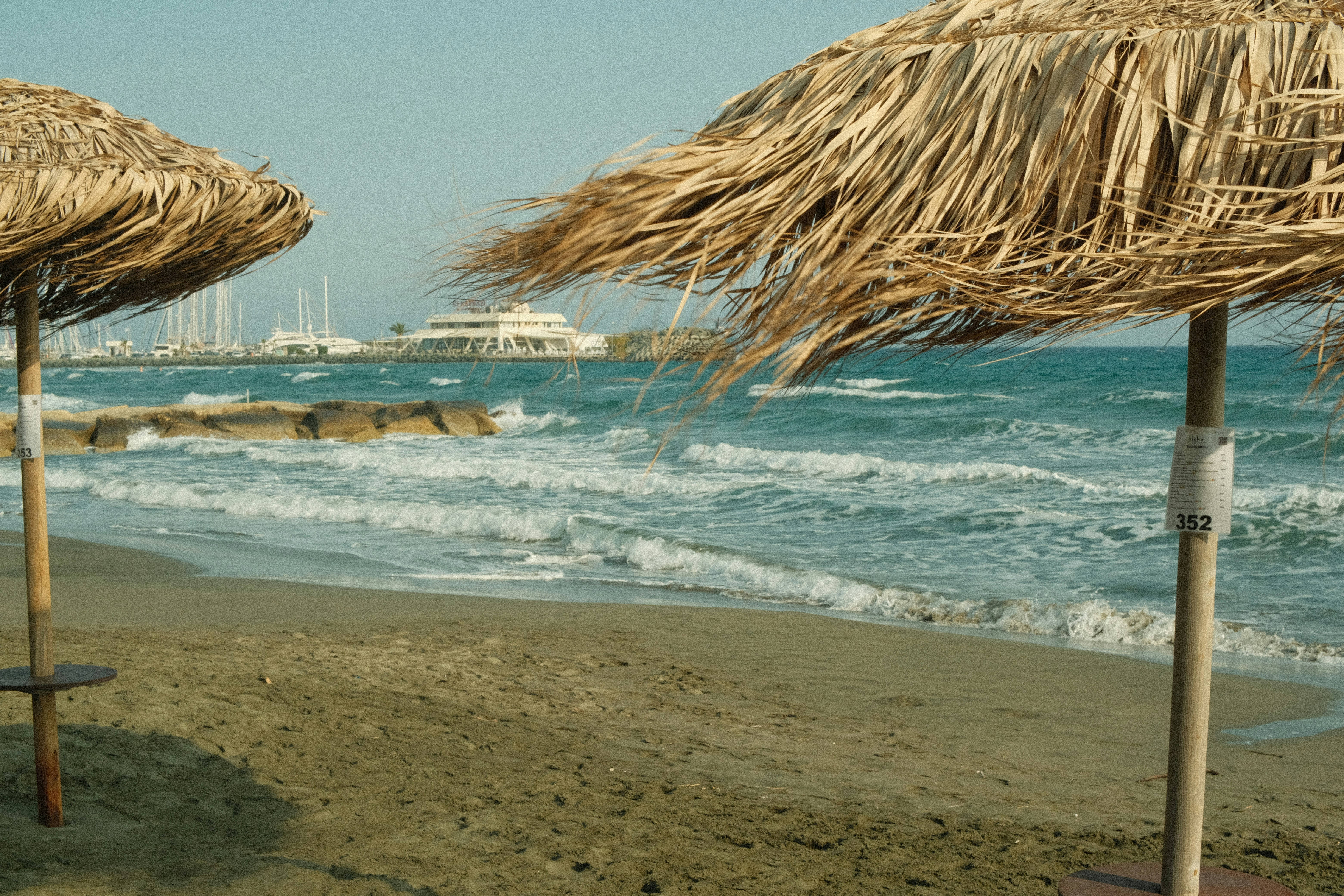 Two thatched umbrellas stand on a sandy beach, overlooking gentle waves and distant boats. The scene evokes a tranquil coastal retreat.