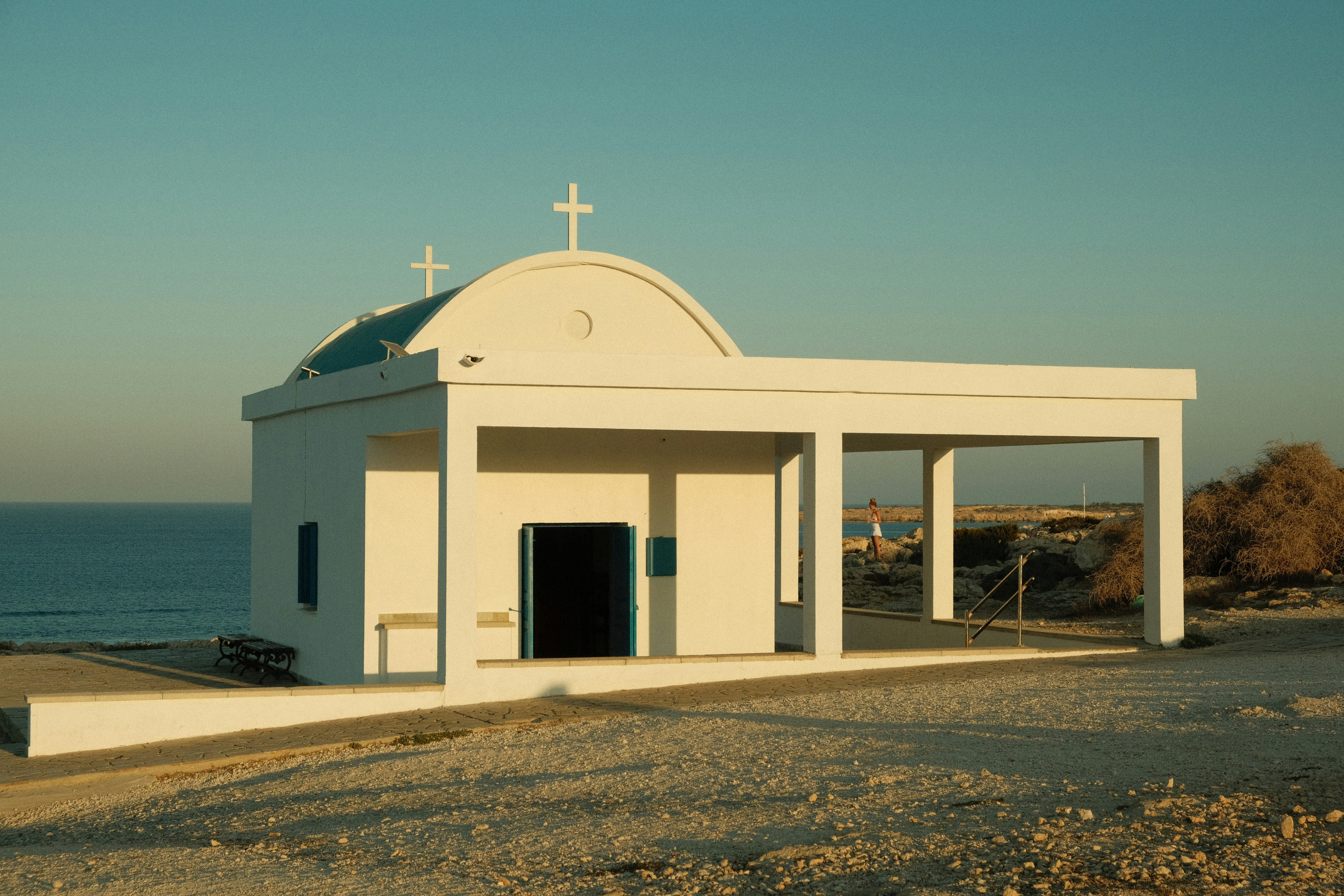 Minimalist white chapel with blue accents overlooking the ocean, framed by golden light of dusk.