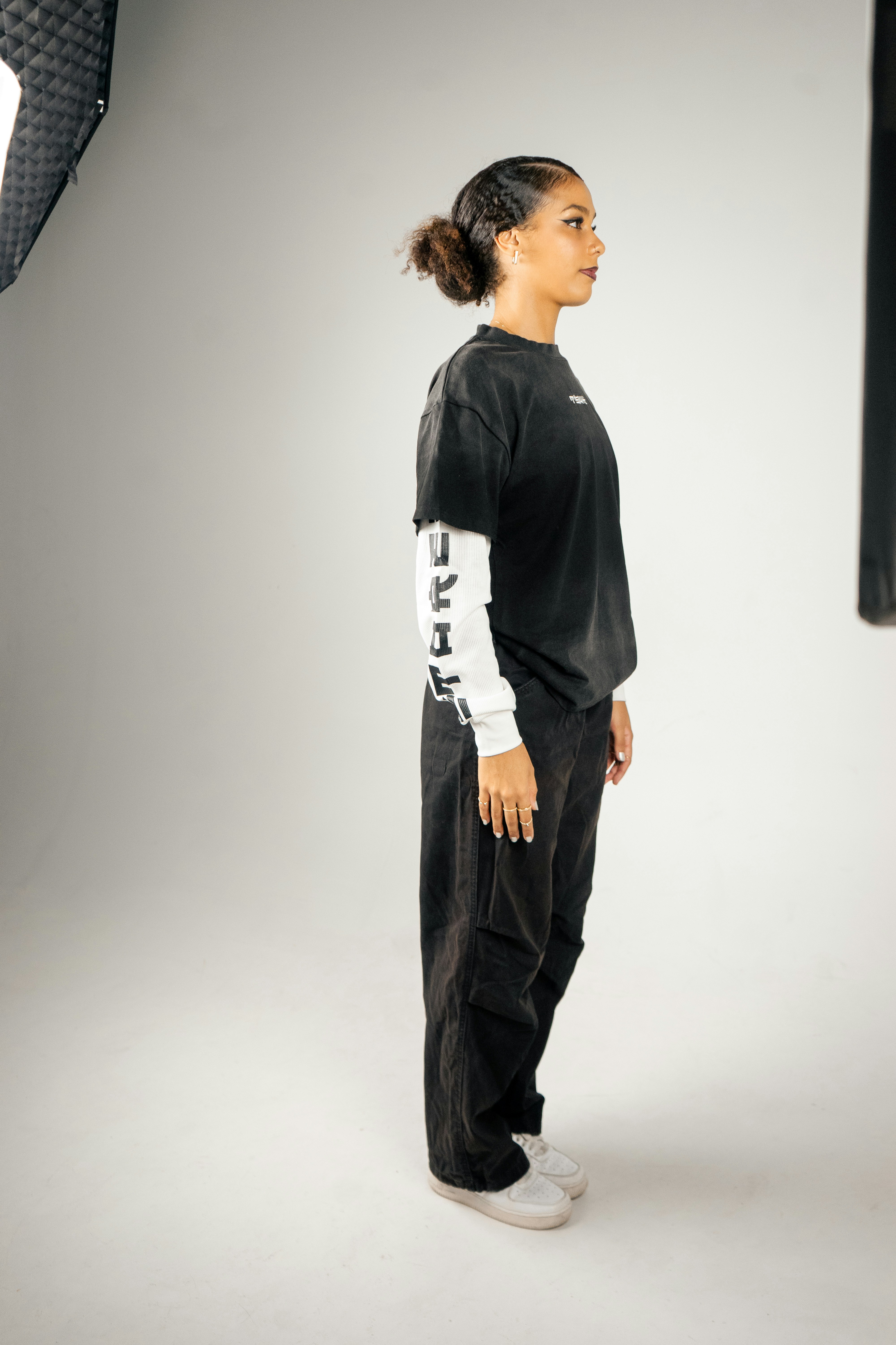 A young woman stands confidently in a black outfit, showcasing a blend of casual and contemporary fashion against a neutral backdrop.