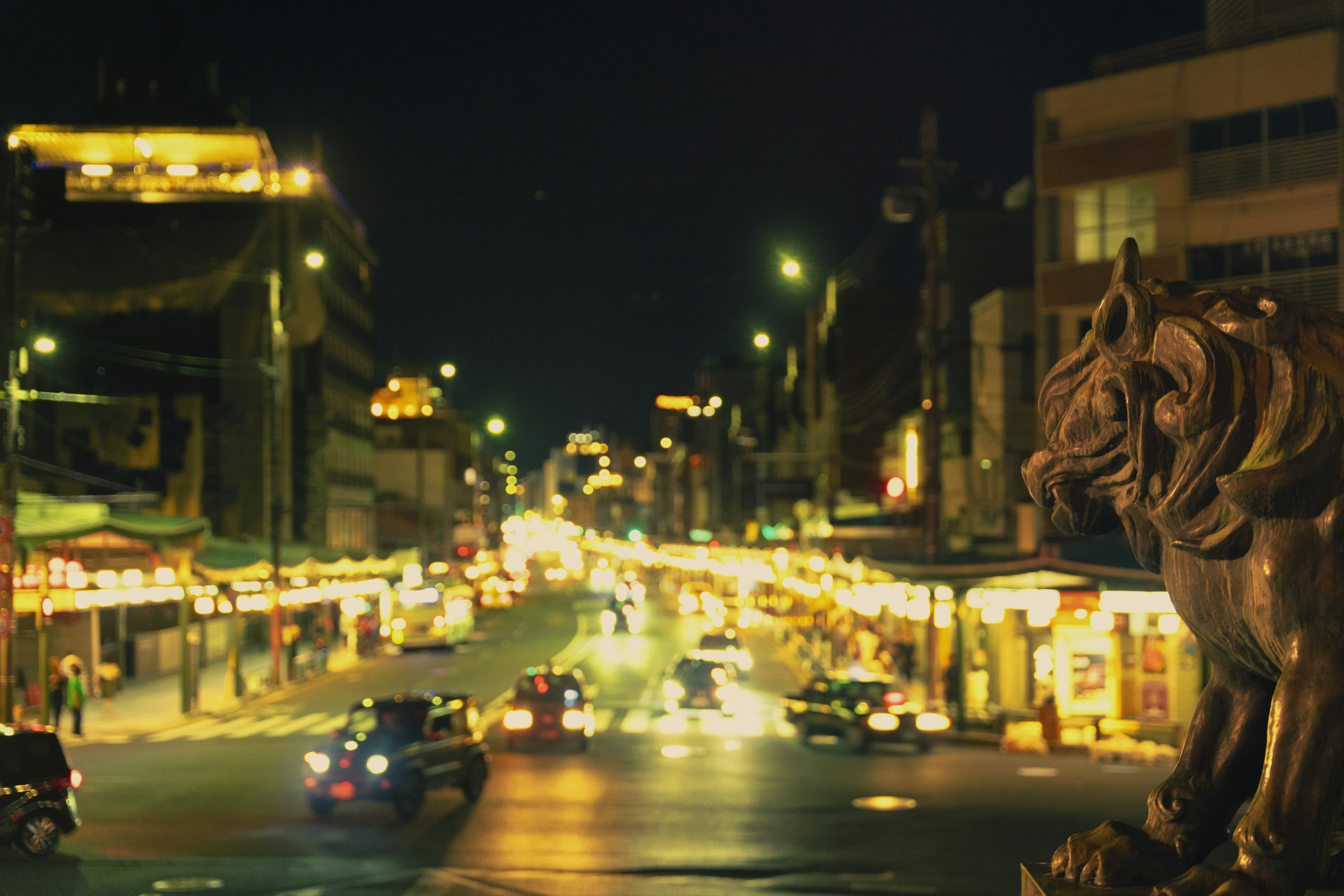 A vibrant city street illuminated at night, featuring a decorative lion statue in the foreground and bustling traffic in the background.