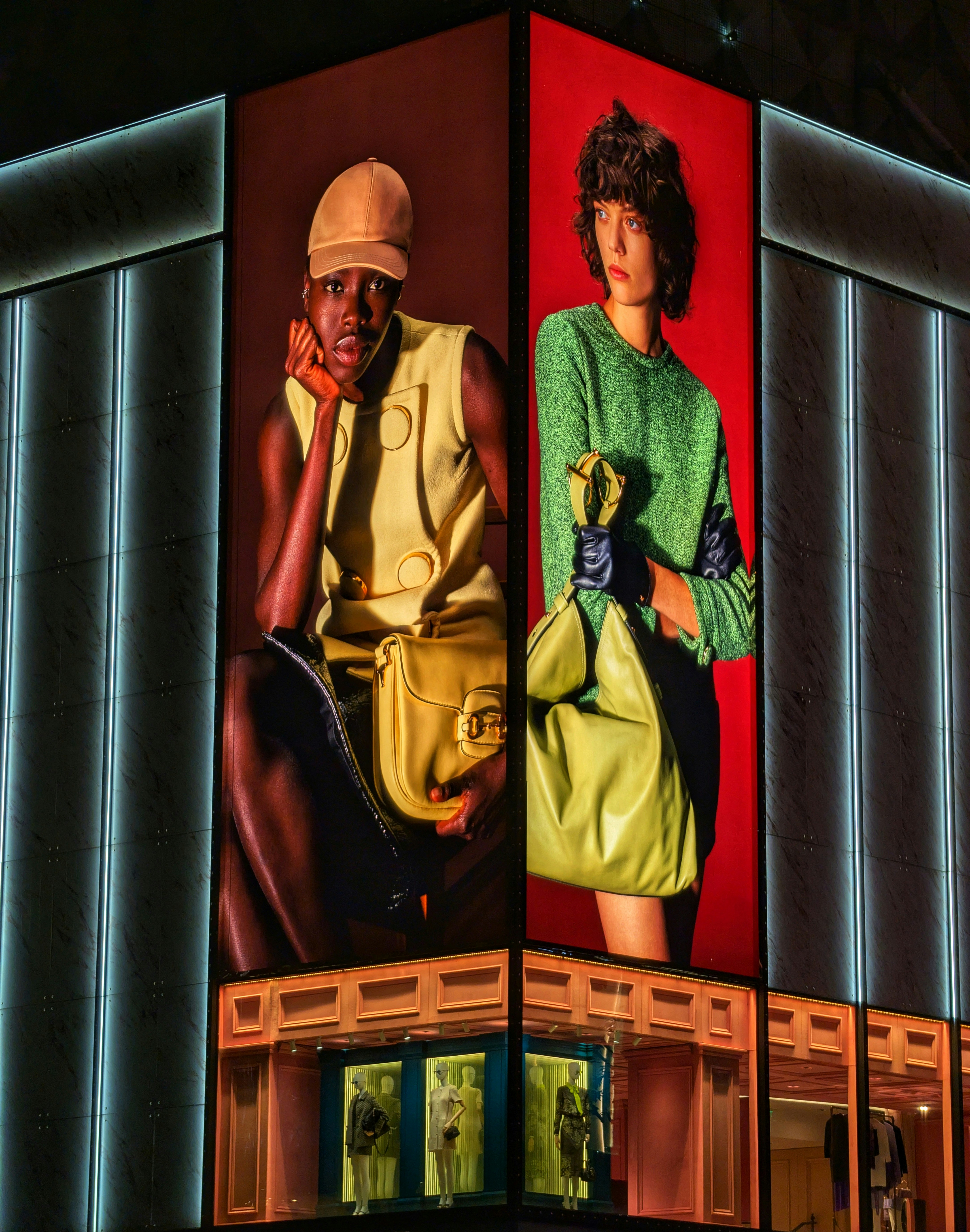 Two fashion models showcased on a building facade, one in a yellow dress and the other in a green sweater, both holding stylish bags against bold backgrounds.