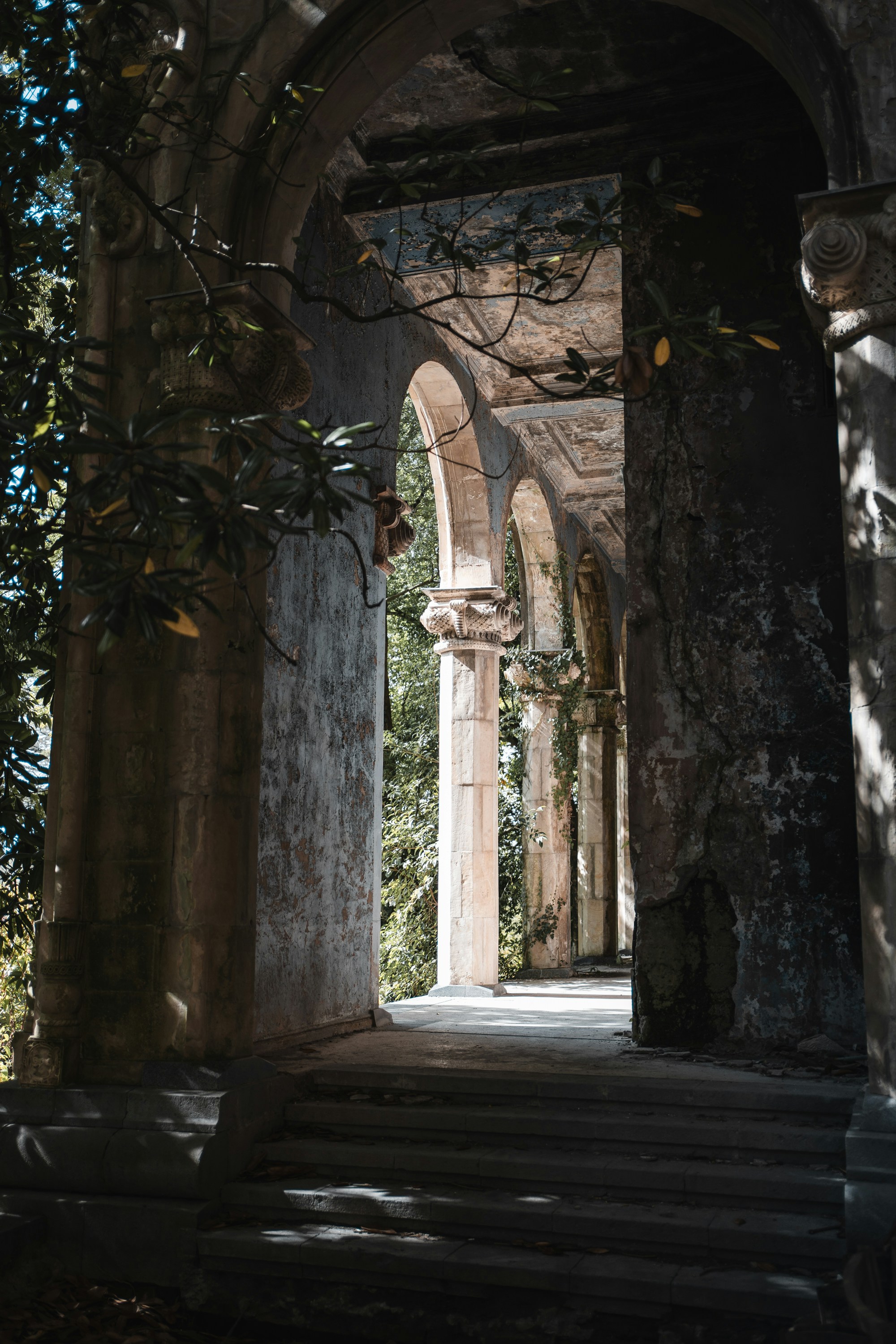 Ancient stone archway framed by lush greenery, showcasing peeling paint and intricate details. Sunlight filters through, creating a serene yet haunting atmosphere.