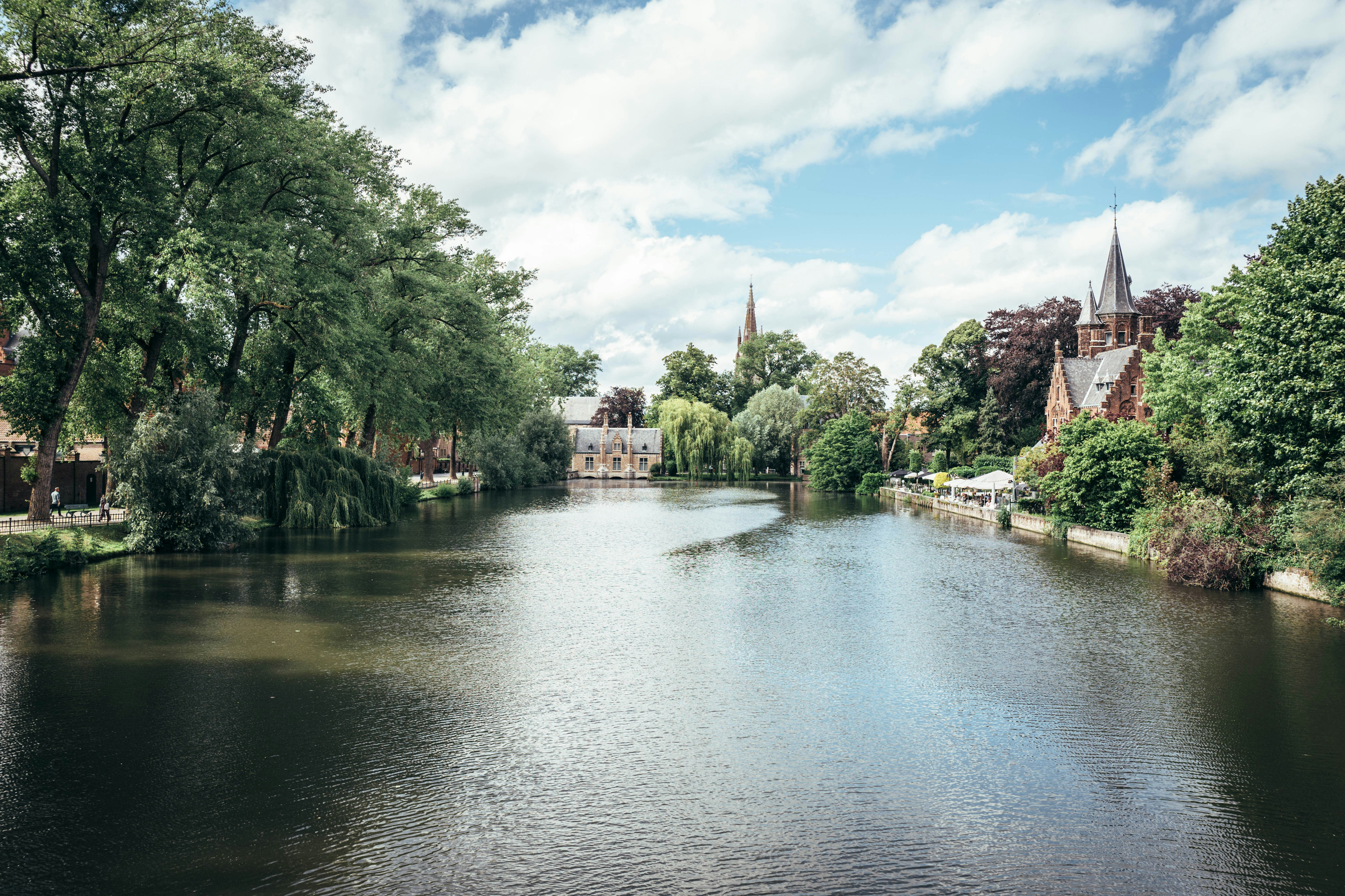 A peaceful riverside scene showcasing lush greenery and historic architecture under a cloudy sky. The calm water reflects the surrounding trees and buildings.