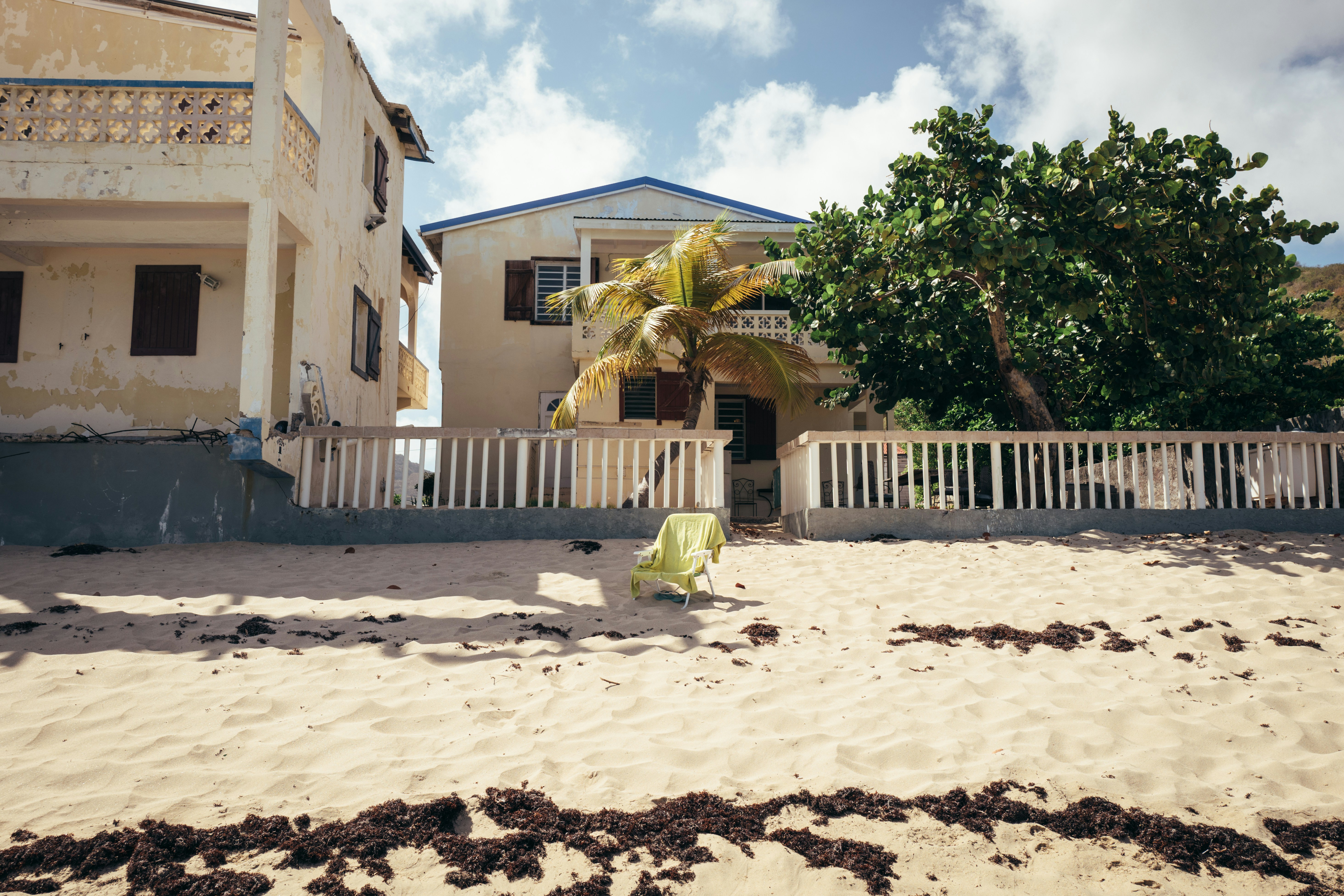 A sunlit beach scene featuring a vibrant green chair on the sand, framed by a charming coastal building and lush vegetation. Relaxation beckons under the warm sun.